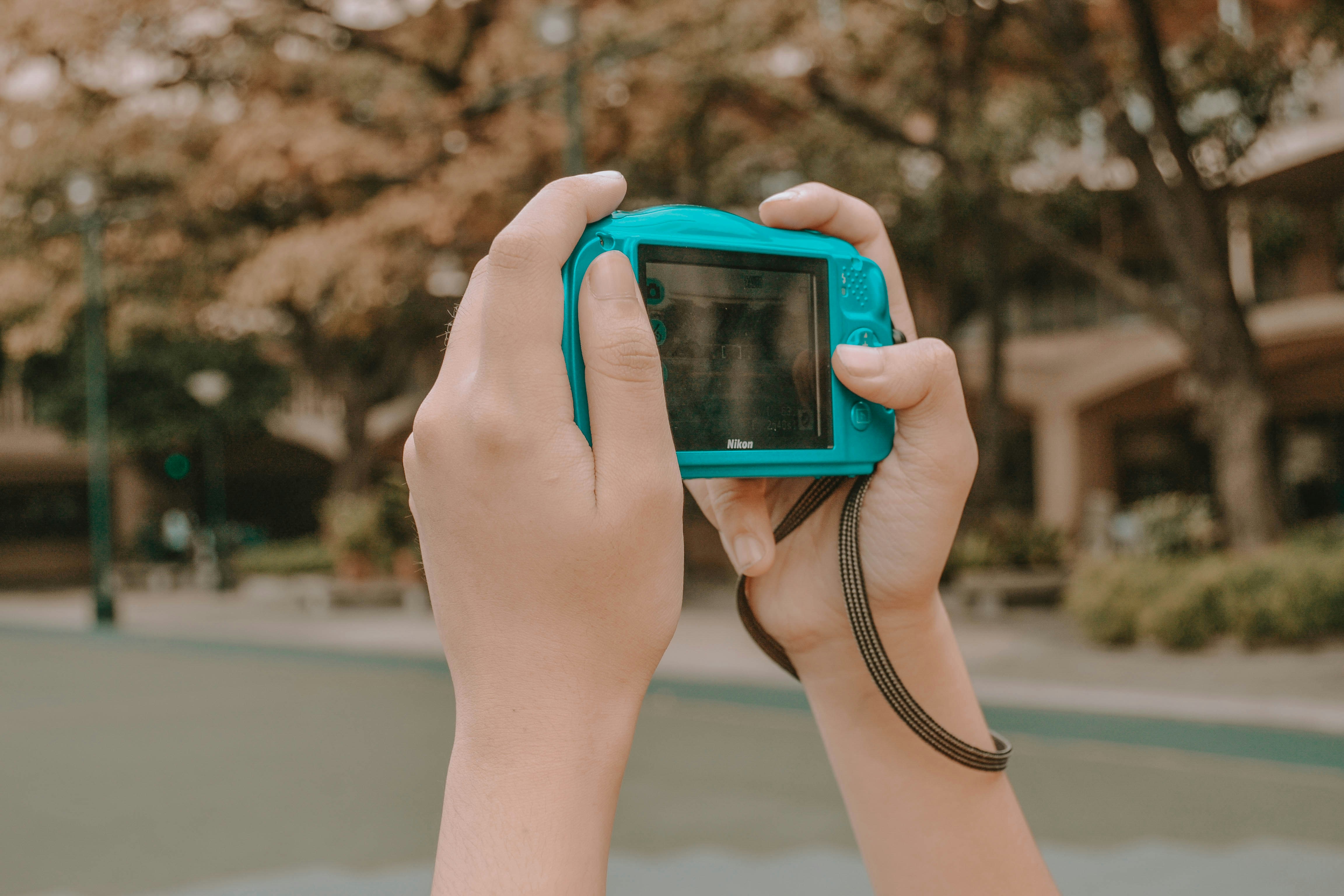 Hands holding a turquoise camera against a backdrop of softly blurred urban foliage. The focus is on the camera, highlighting its unique color and design.
