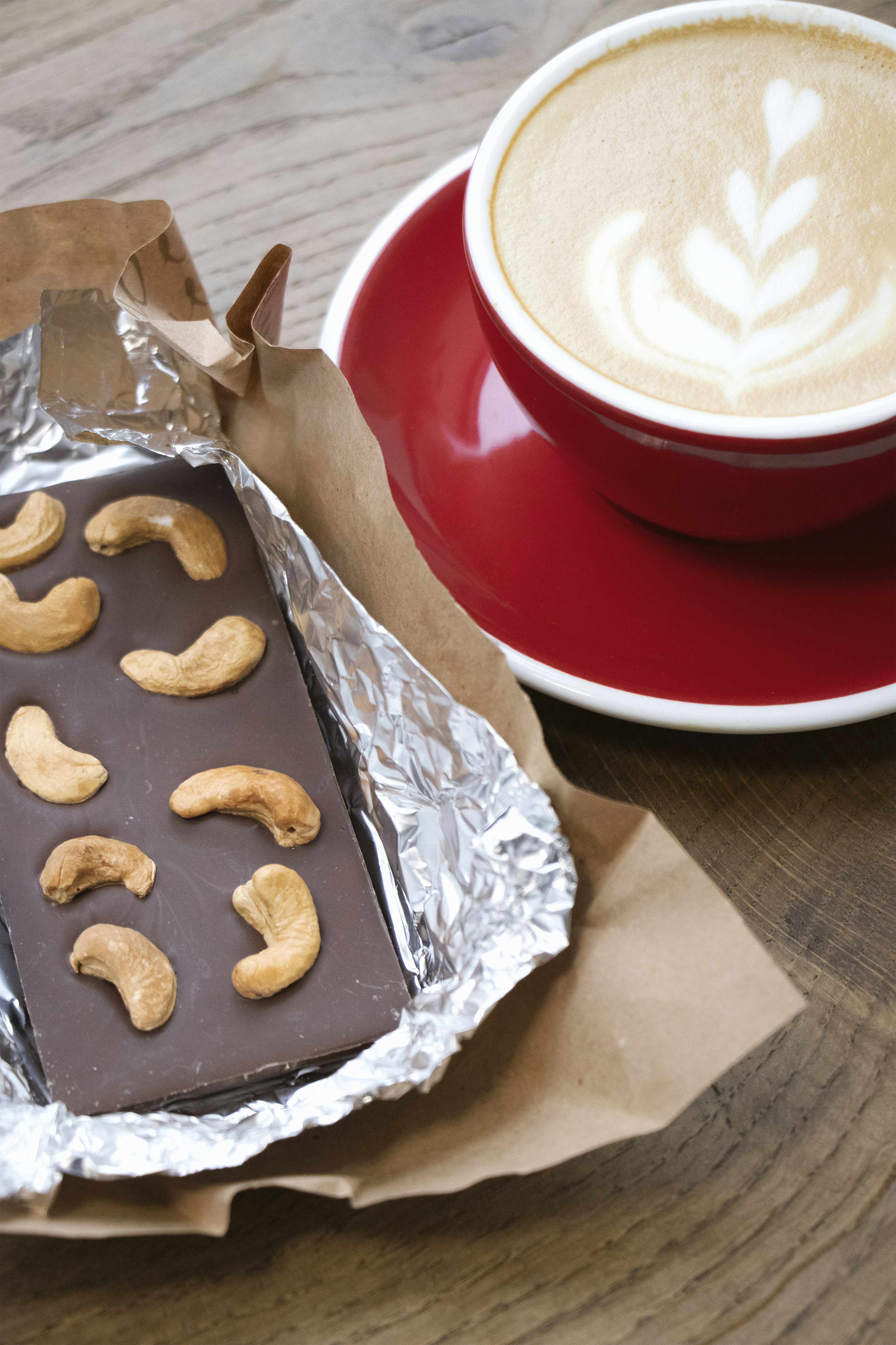A rich chocolate bar topped with cashews rests on crinkled foil beside a beautifully crafted latte in a red cup. 