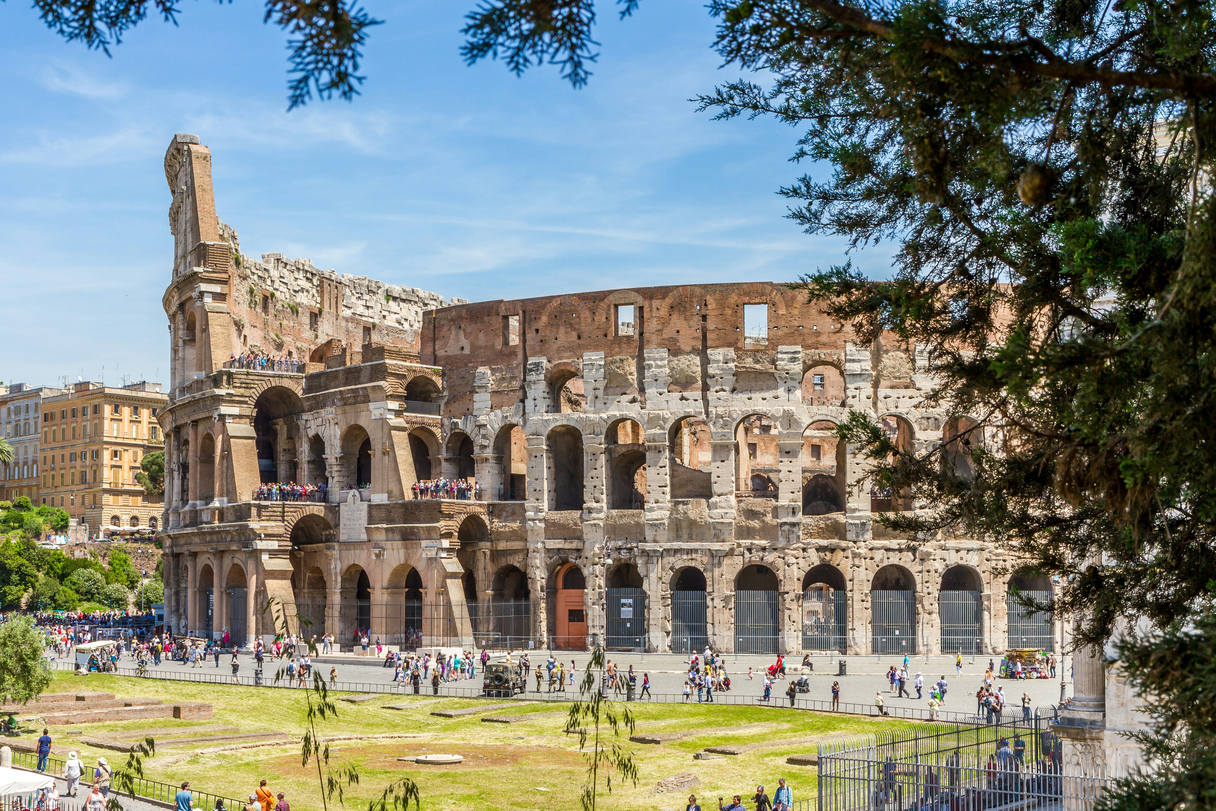The ancient Colosseum stands amidst bustling crowds and vibrant greenery under a clear blue sky.