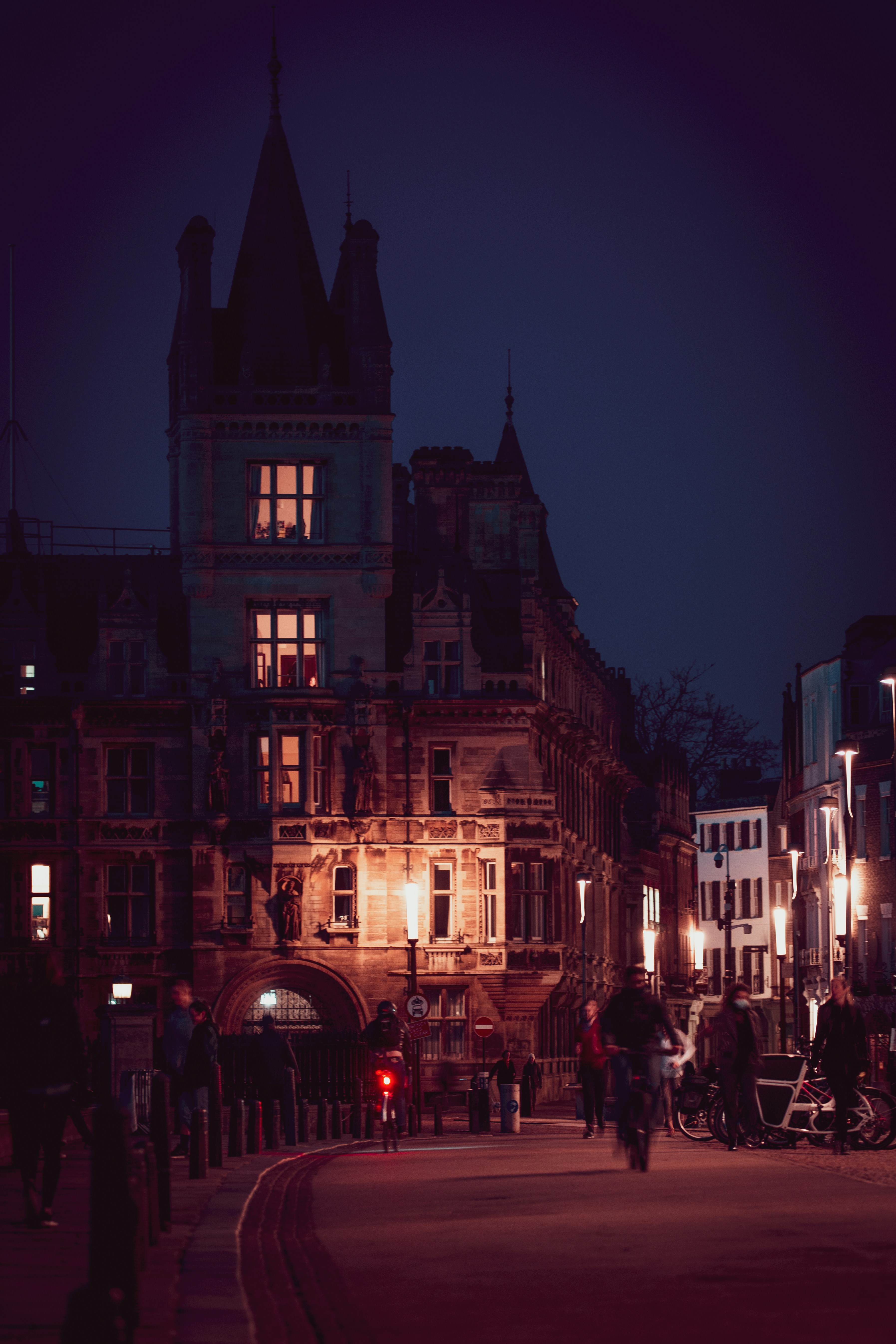 Illuminated Victorian building at dusk, with pedestrians and bicycles along a cobblestone path.