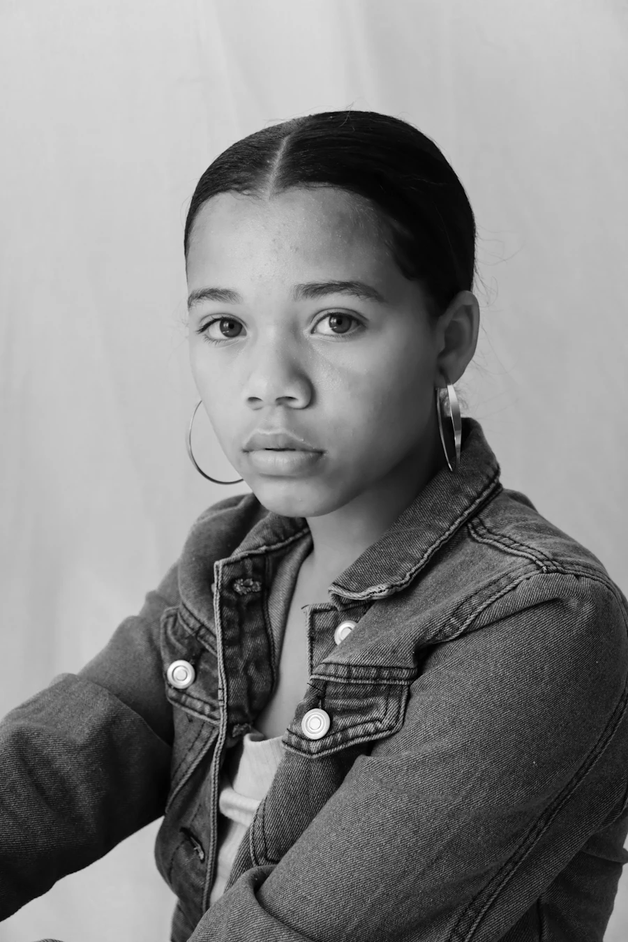 Black-and-white portrait of a young woman with hoop earrings and a denim jacket, facing the camera against a plain backdrop.