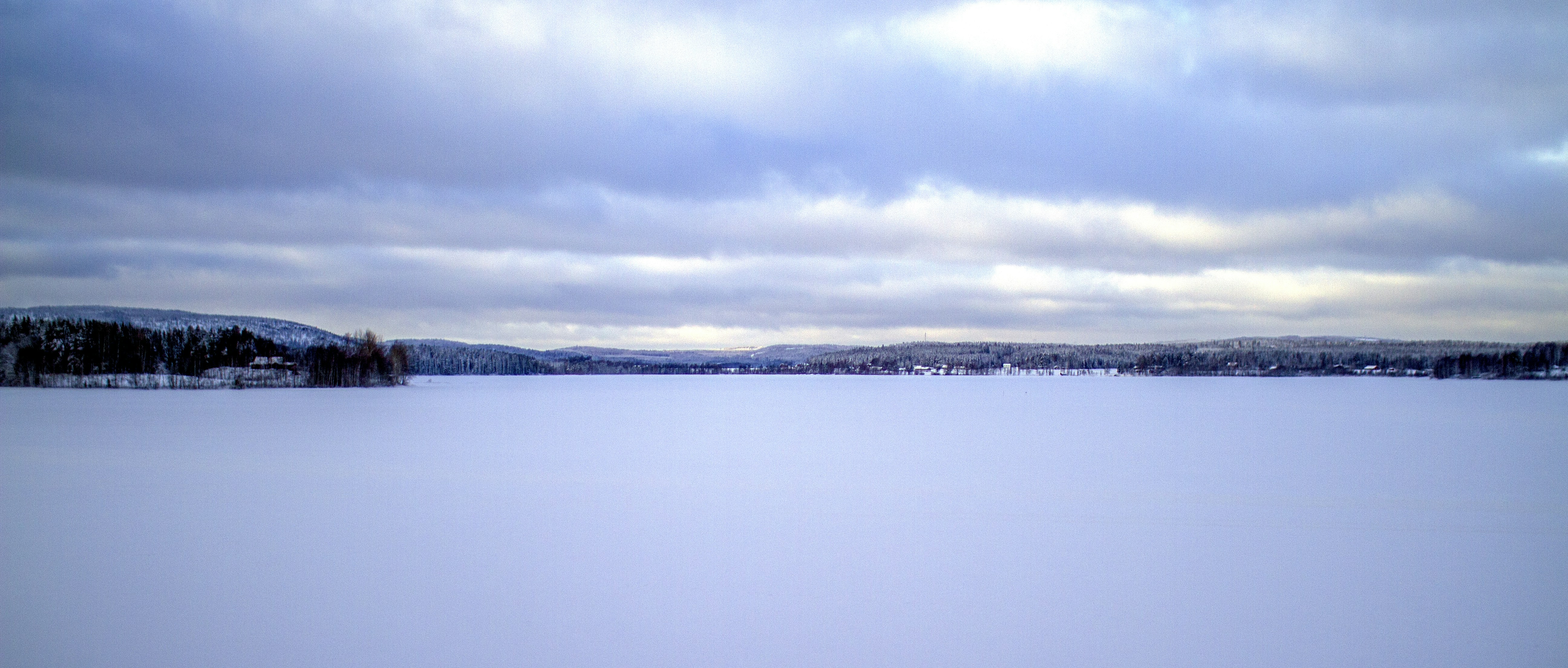 Expansive frozen lake under a soft, cloudy sky, evoking a serene winter landscape. The scene captures the stillness and tranquility of a snowy environment.