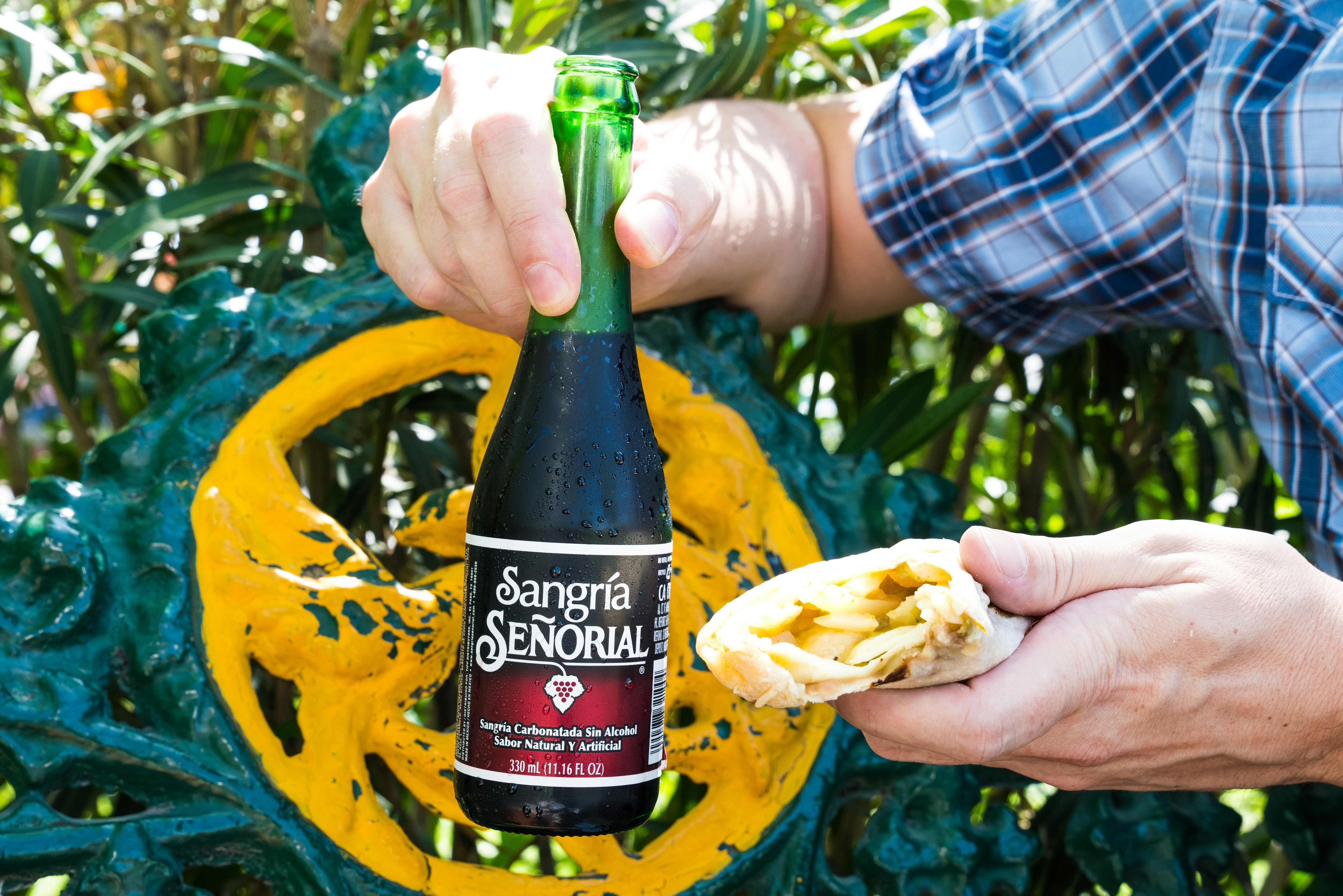 A hand holds a small dark bottle labeled Sangría Senorial next to a folded tortilla in front of a textured, colorful garden sculpture. This image emphasizes a casual outdoor snack moment with bold label details.
