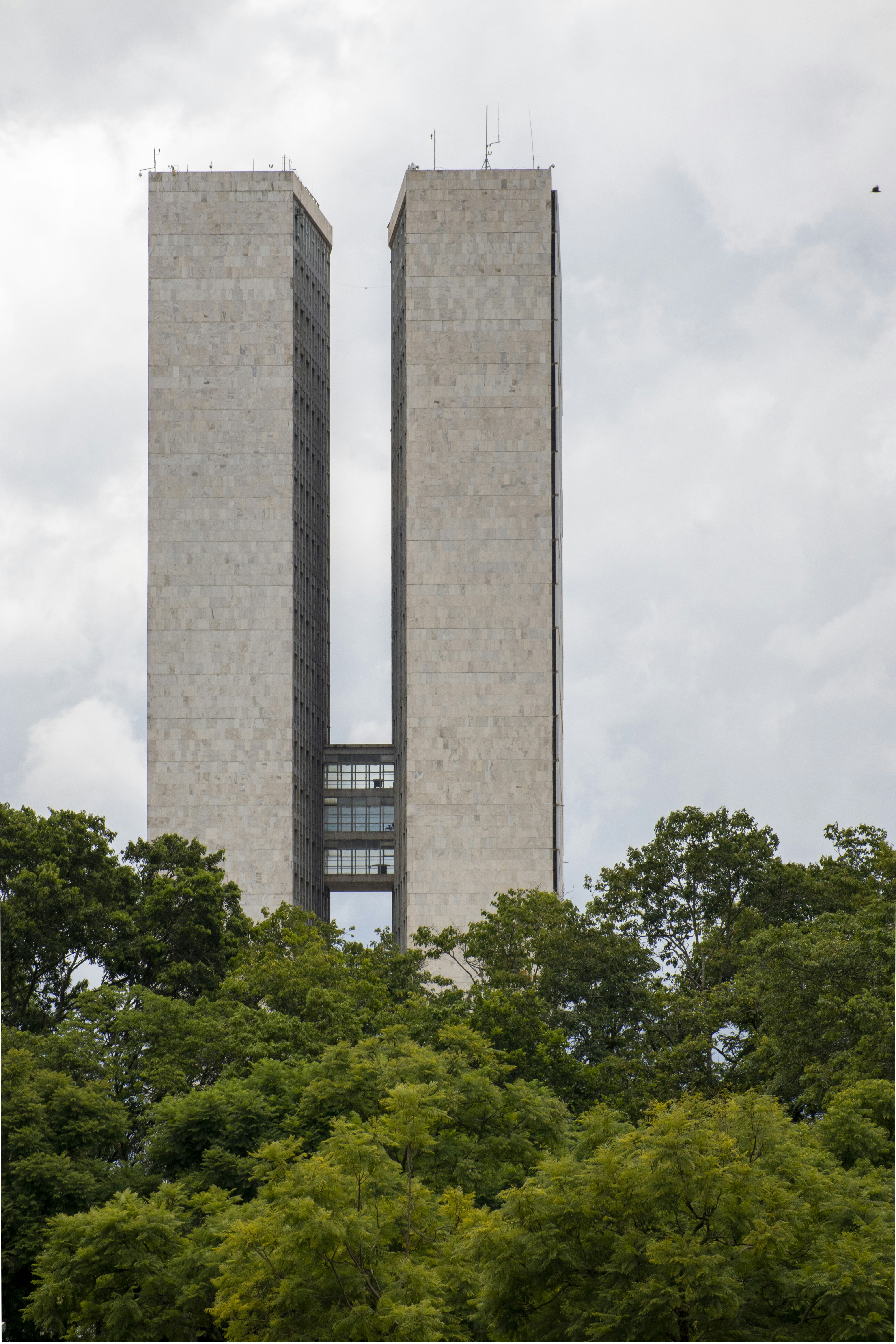 Two tall, rectangular towers rise above lush green foliage under a cloudy sky. The modern architecture contrasts with the surrounding trees.