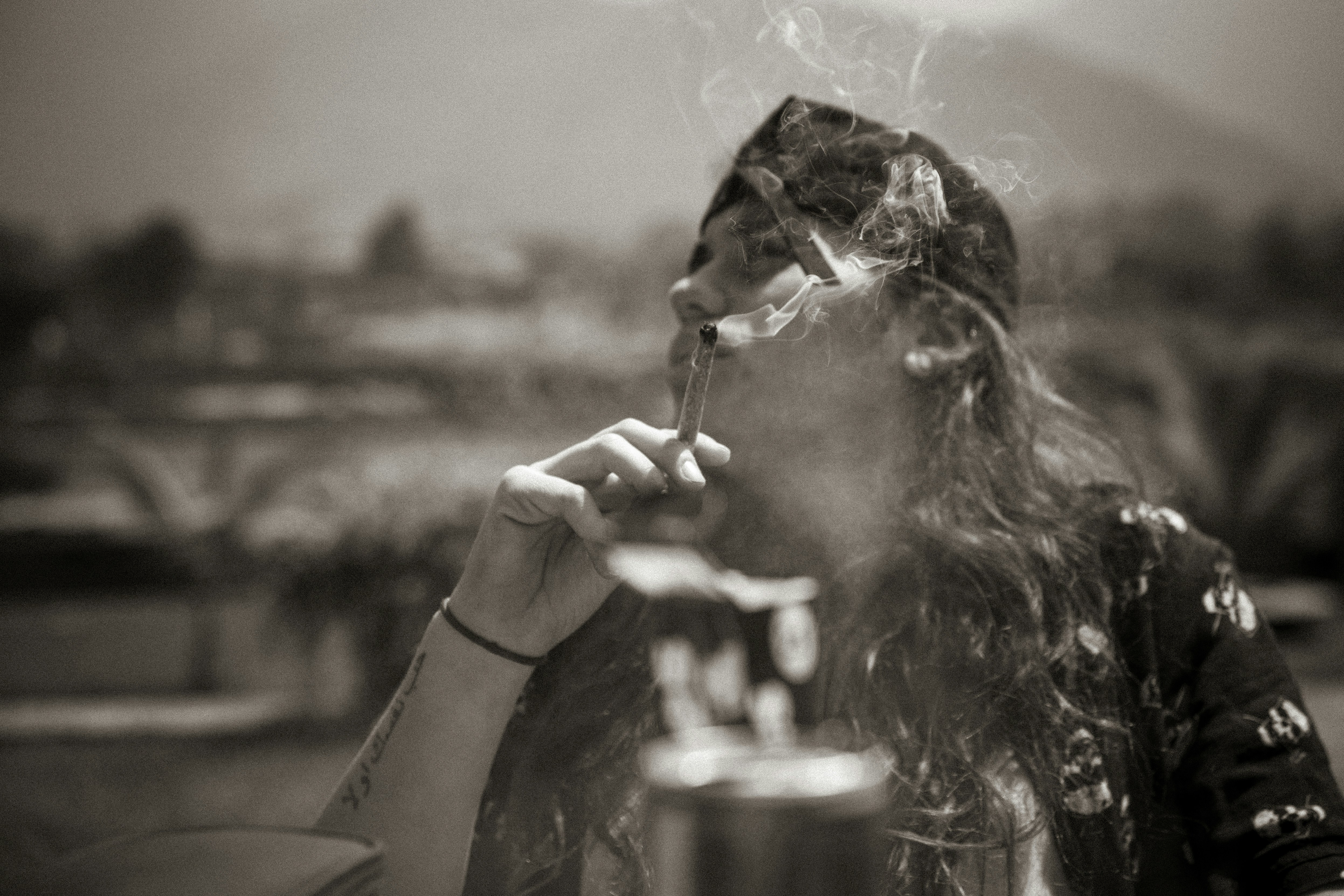 A person with long hair exhales smoke while lost in thought, surrounded by a blurred outdoor backdrop. The black-and-white tone enhances the mood of reflection.
