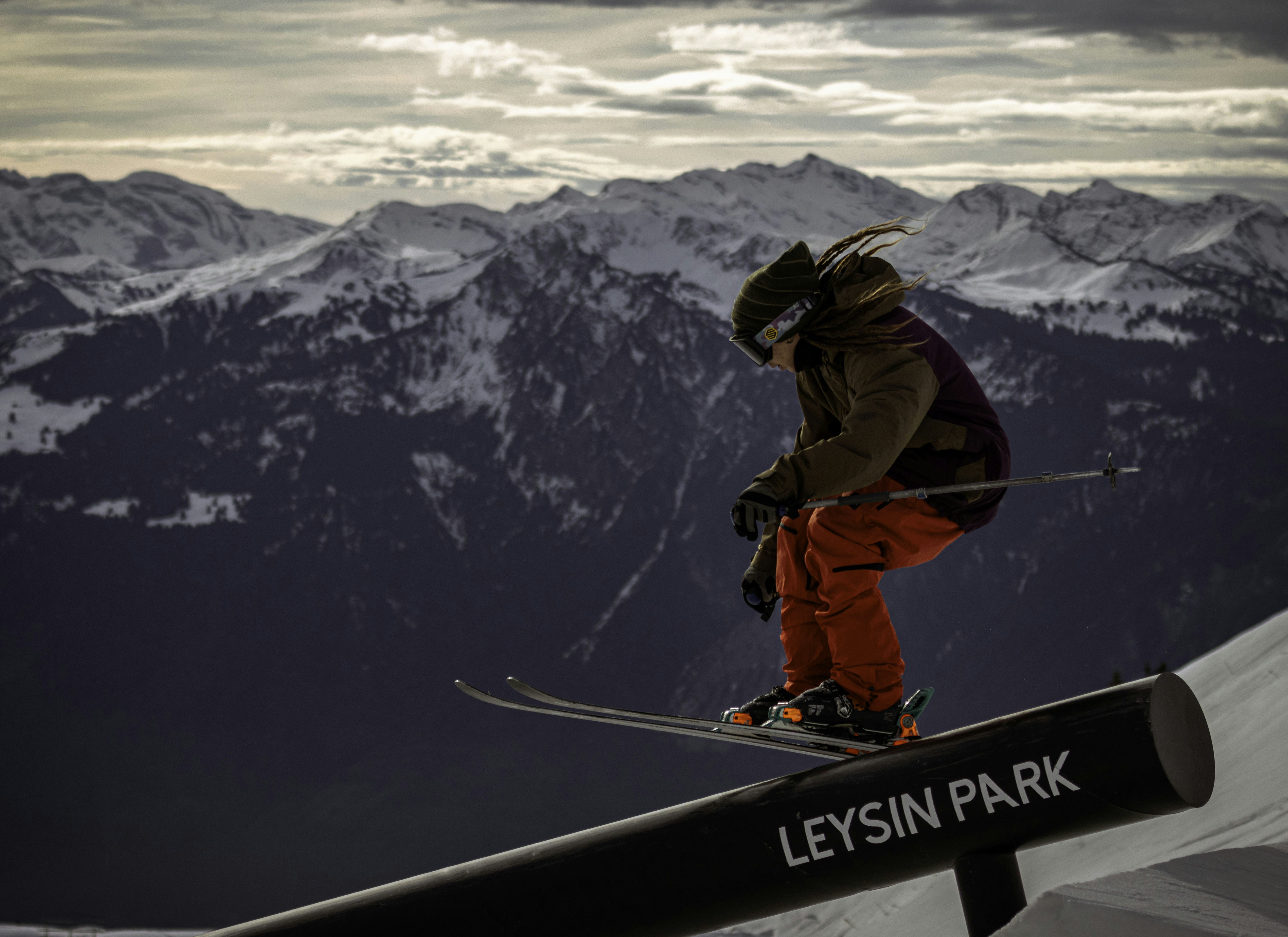 Skier performing a trick on a rail at Leysin Park, surrounded by majestic snow-capped mountains under a dramatic sky.