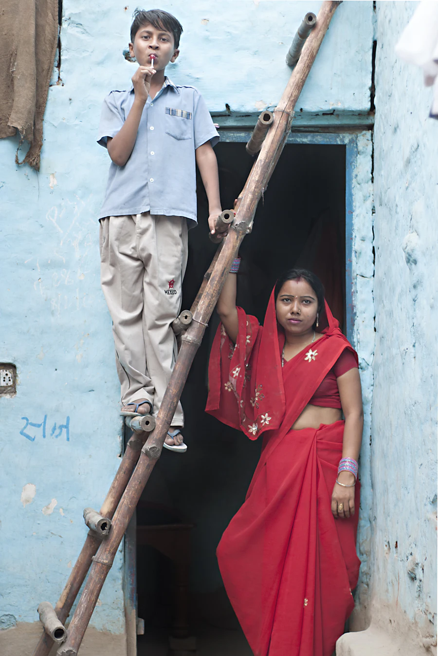 Boy standing on a bamboo ladder at a blue doorway while a woman in a red sari poses below him. The narrow entryway and weathered wall create a vivid neighborhood portrait.
