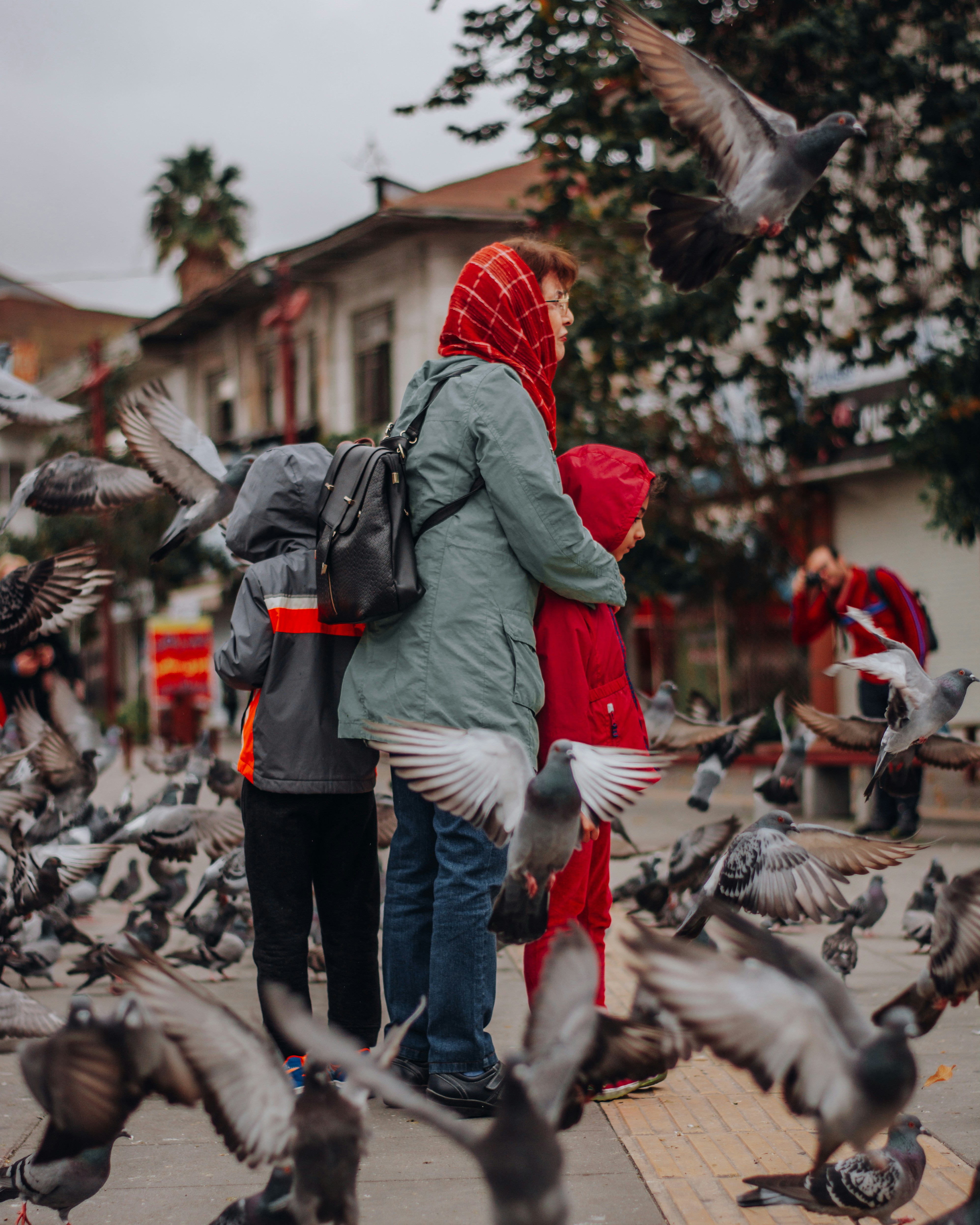A woman embraces two children amidst a flurry of pigeons in a bustling urban setting, capturing a moment of joy and connection.
