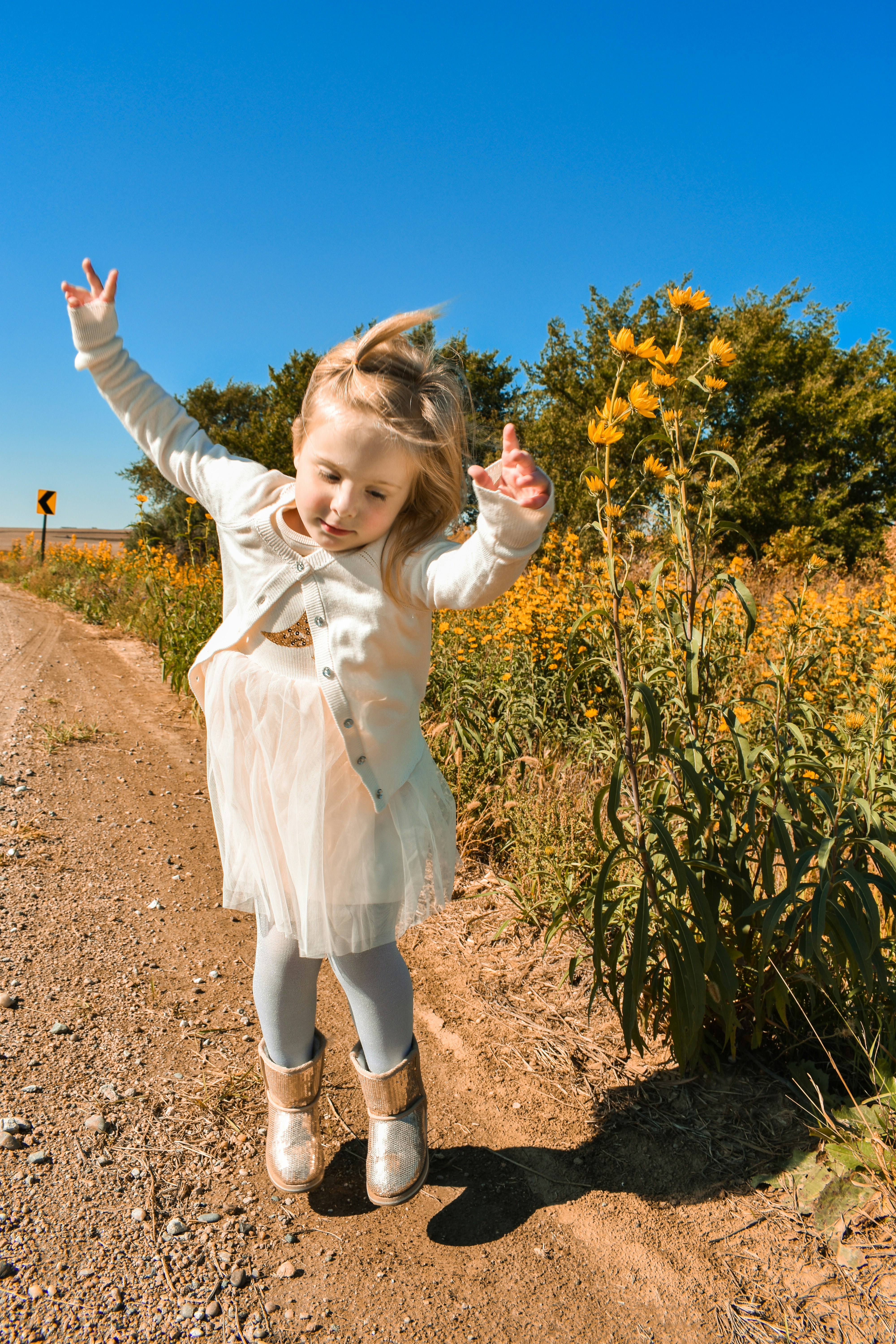 A young girl in a white dress joyfully dancing along a dirt path surrounded by vibrant yellow flowers and greenery. The clear blue sky enhances the cheerful atmosphere.