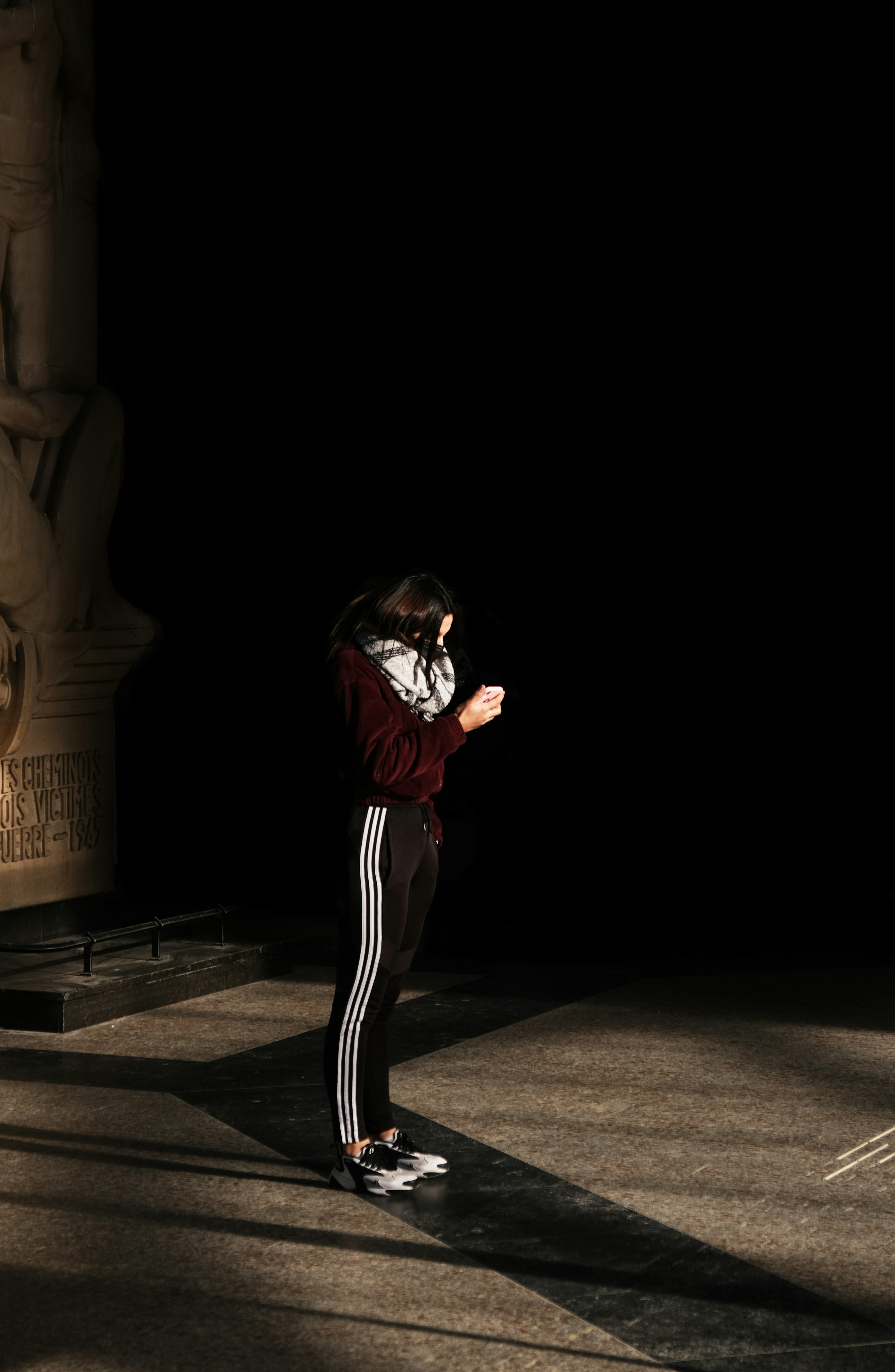 A solitary figure stands in a darkened environment, engrossed in her phone, with a faint beam of light highlighting her presence.