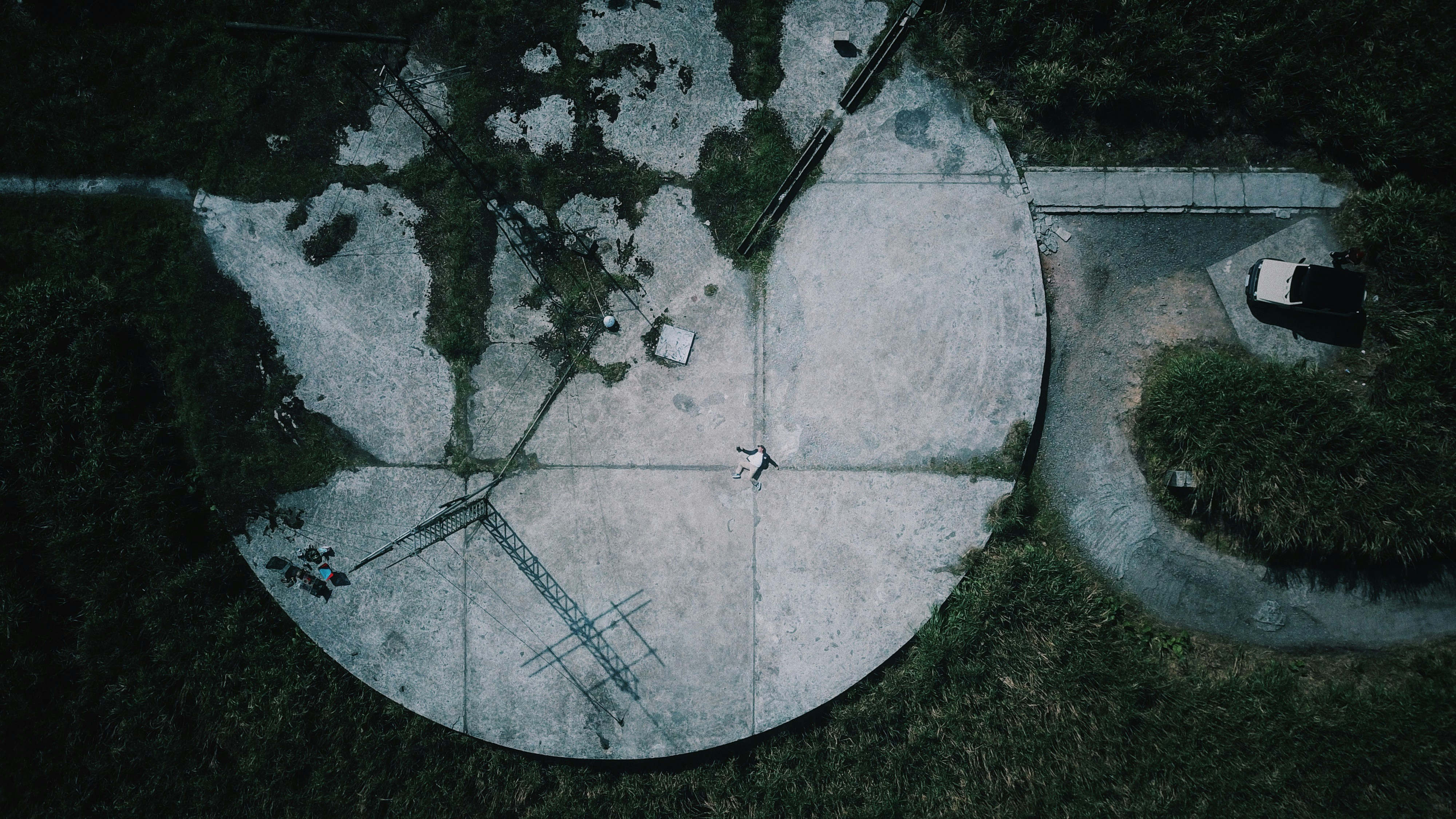 Aerial view of a circular, weathered concrete structure surrounded by overgrown grass and a winding path, conveying a sense of neglect and time passing.