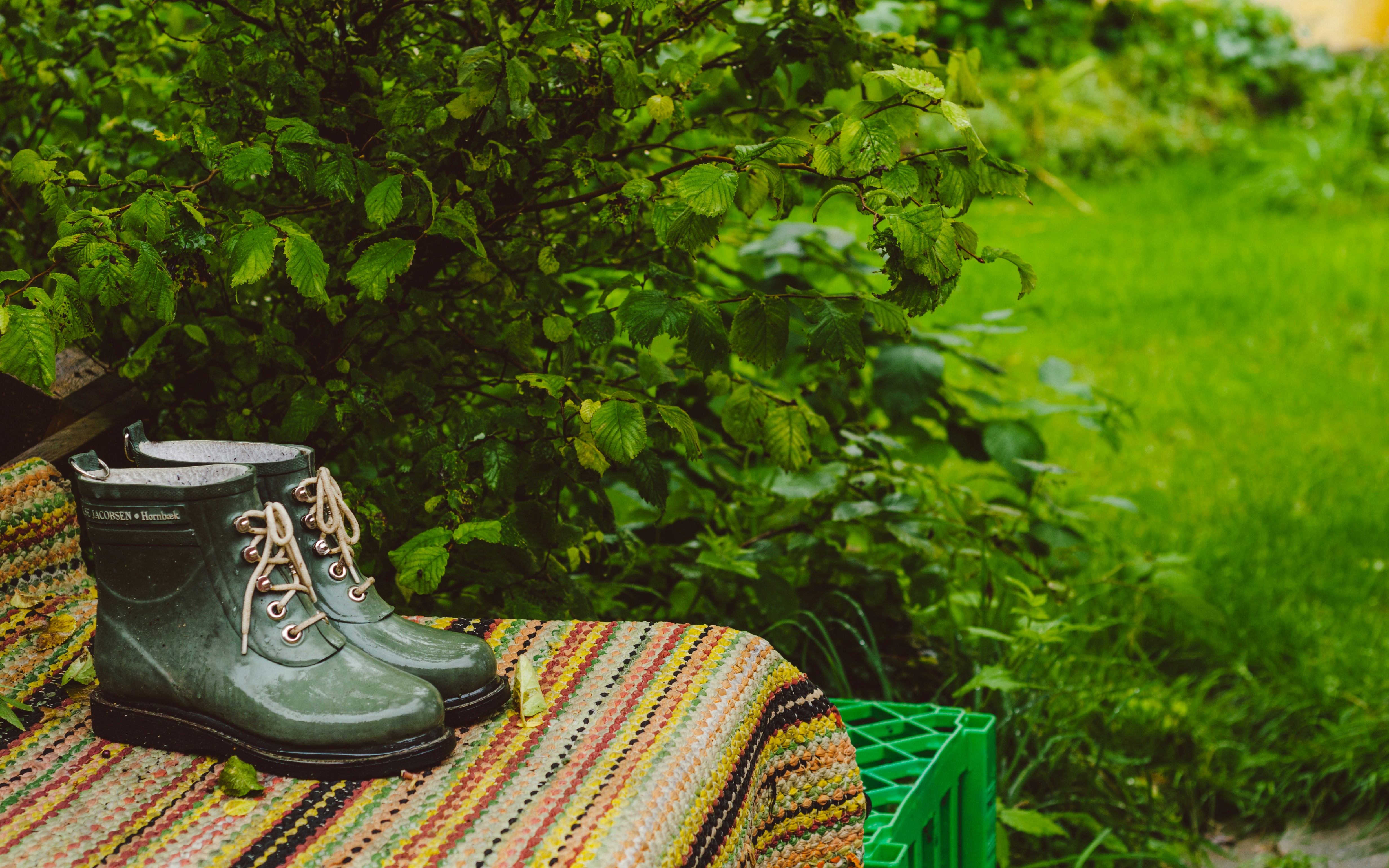 Green hiking boots resting on a woven striped mat beside a verdant garden