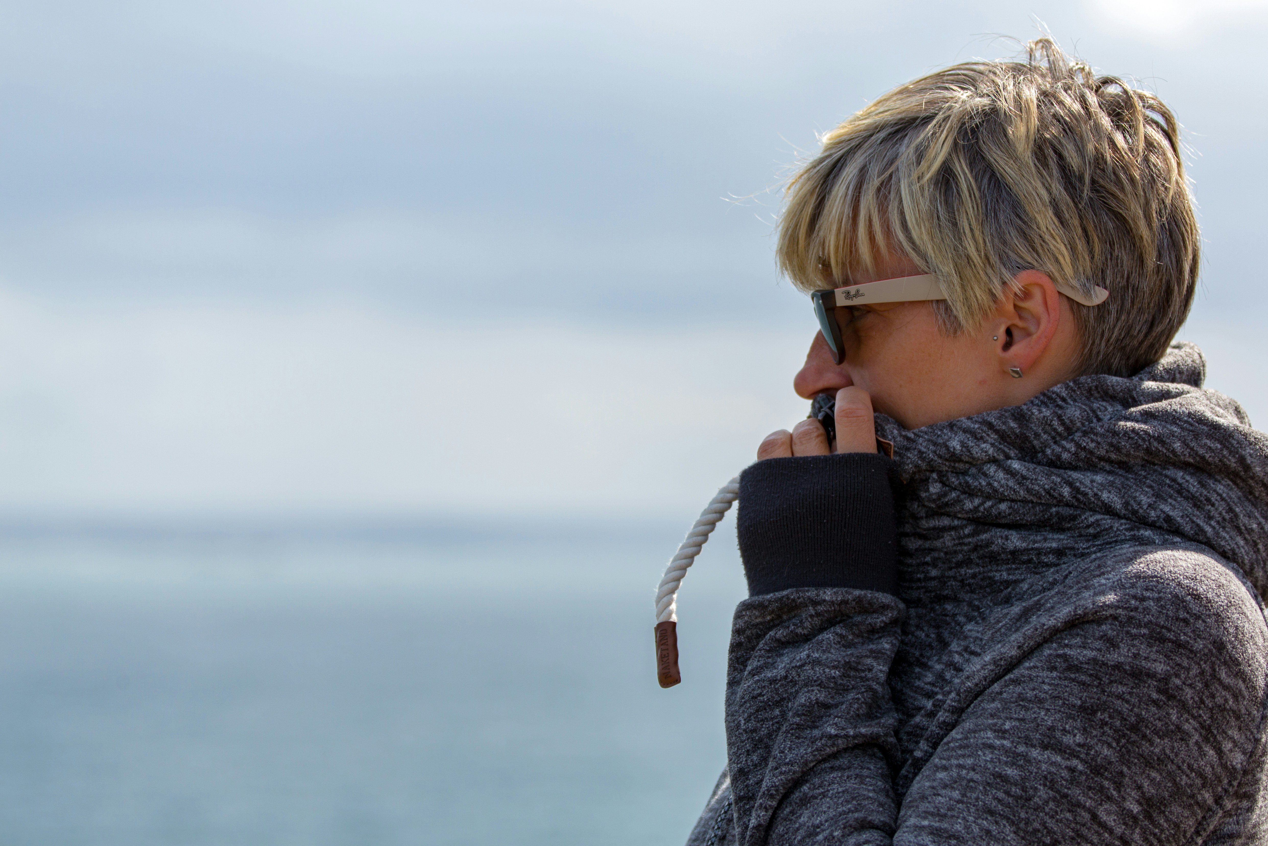 A woman in a gray hoodie stands pensively, gazing towards the ocean, with a soft breeze tousling her hair.