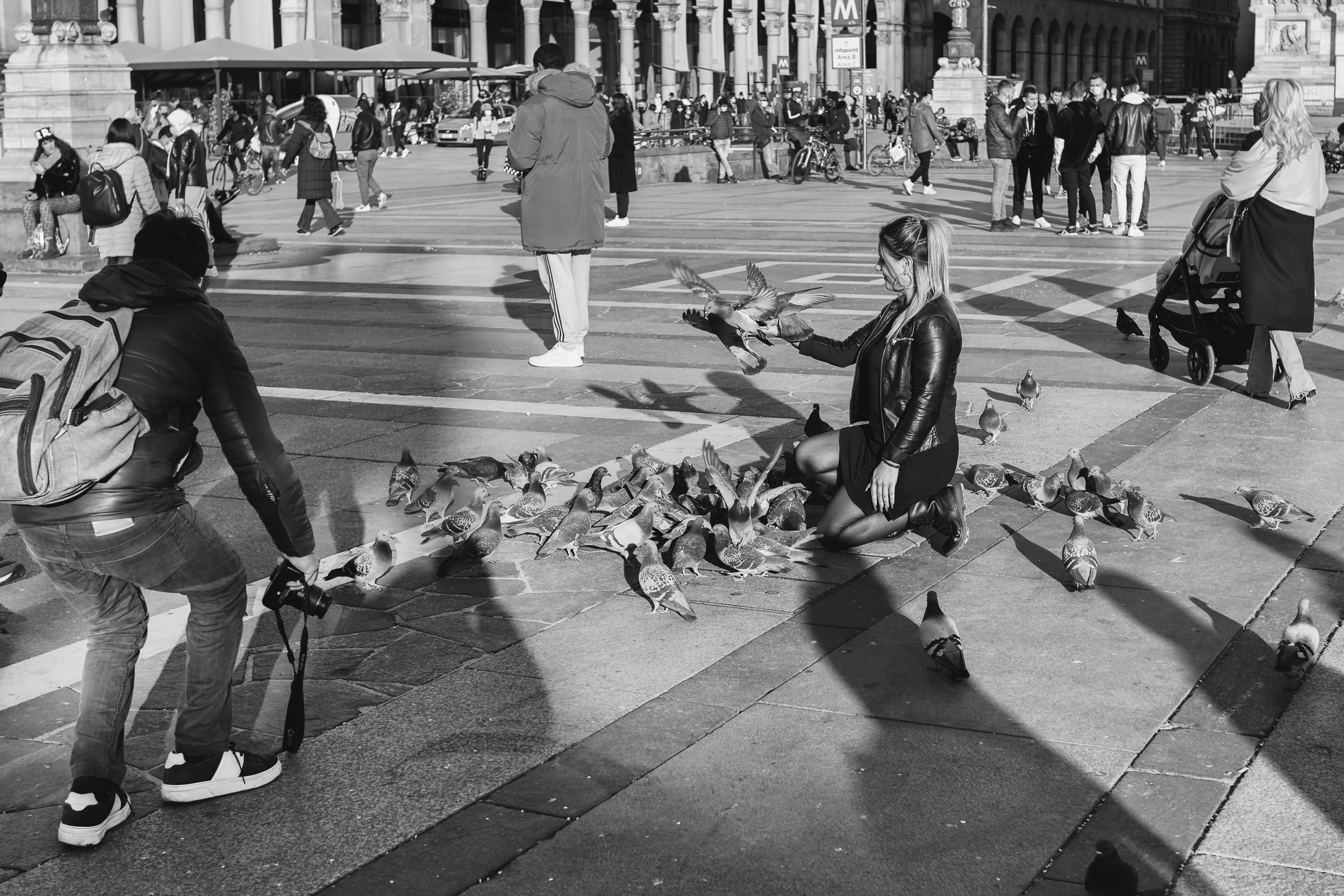 Grayscale street scene of a woman seated on a city plaza feeding pigeons. A photographer to the left captures the moment as pedestrians pass by.