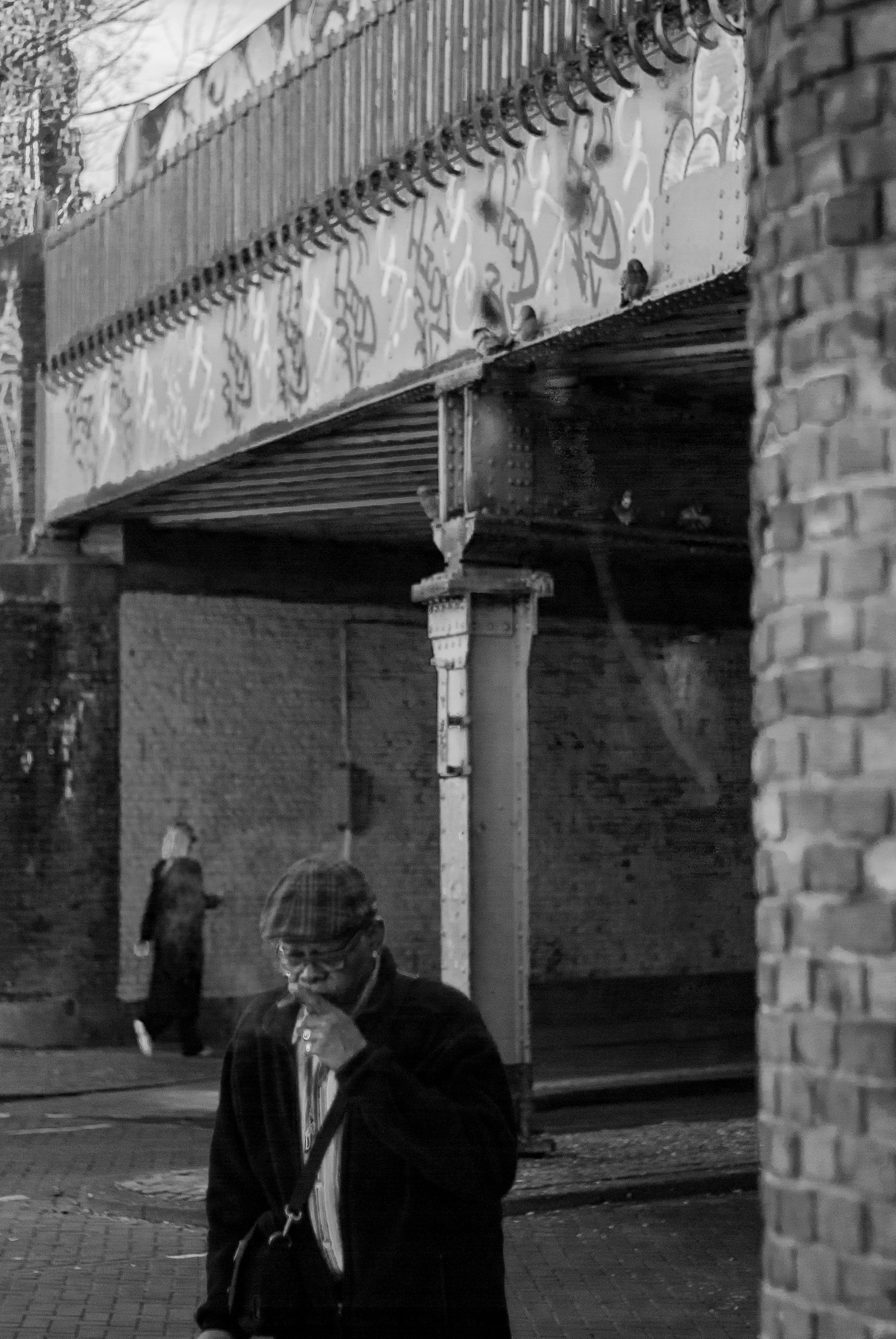 An elderly man pauses to smoke beneath a graffiti-covered bridge, while a figure walks in the background, capturing the essence of urban life and solitude.