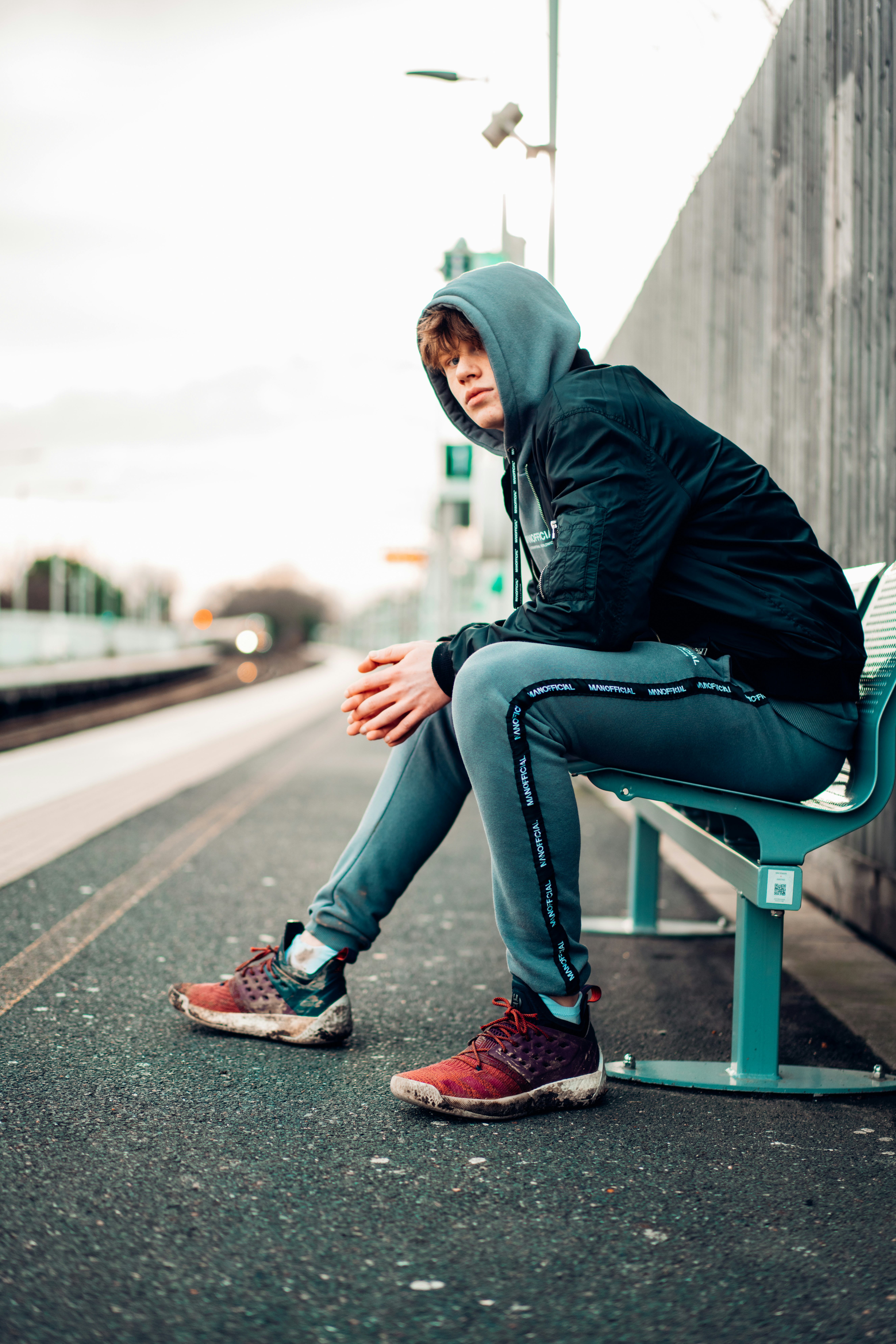 Young man in a hoodie sitting on a bench at a train station, lost in thought as the world moves around him.