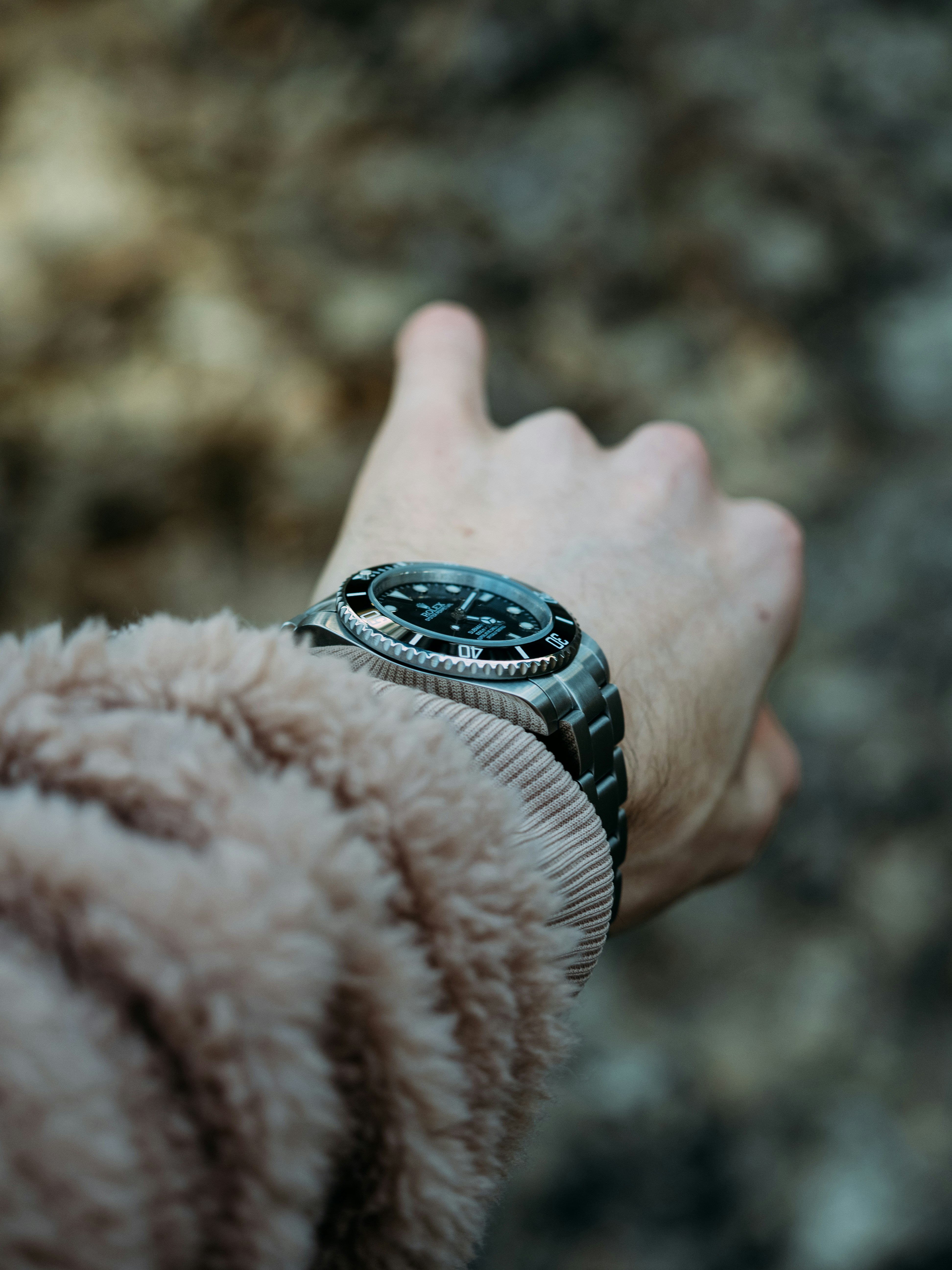 A hand extends forward, showcasing a sleek watch against a blurred natural backdrop. The focus highlights the intricate details of the watch's face.