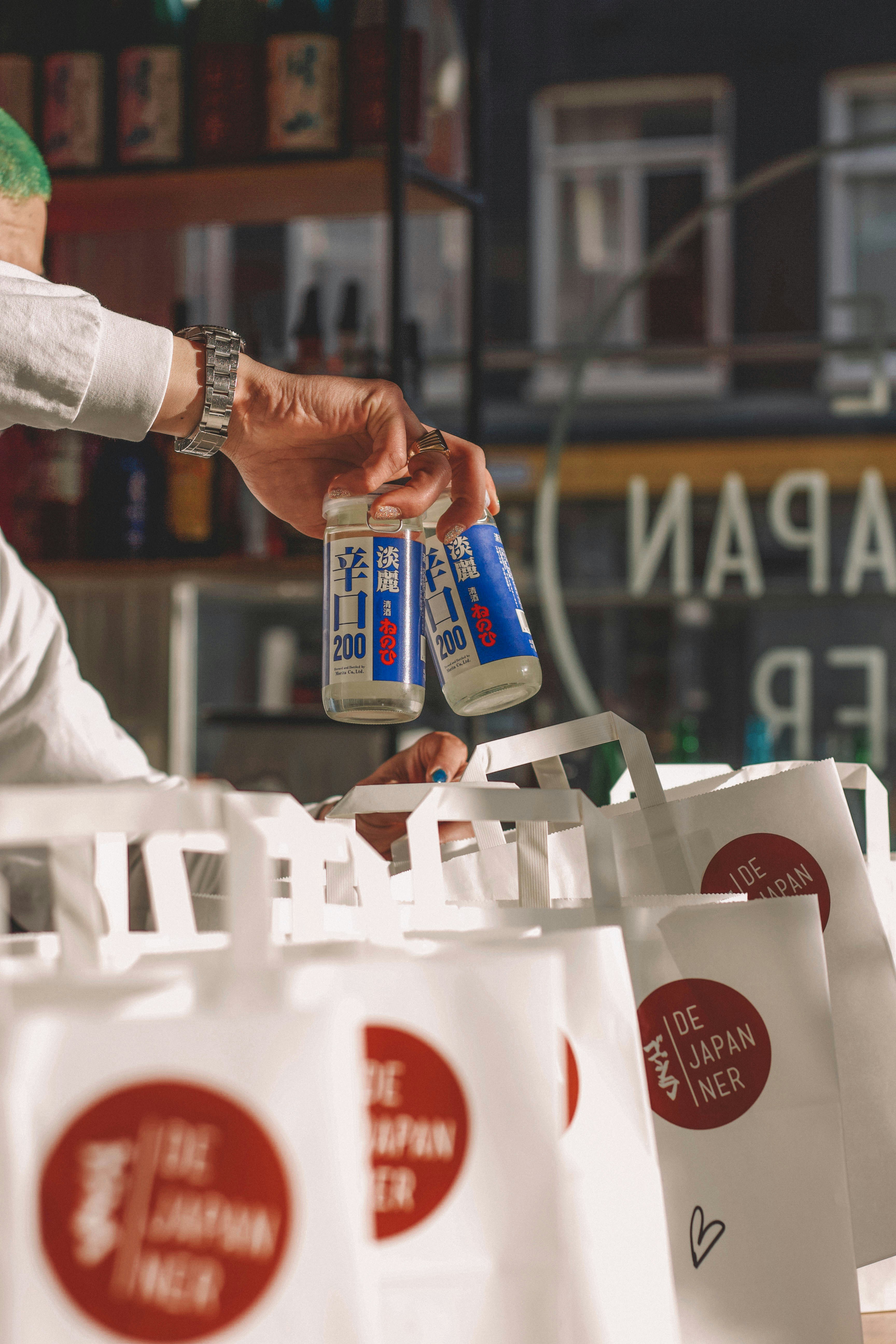 Two Blue-Labeled Sake Bottles Hover Over White Gift Bags in a Quiet Bar Scene