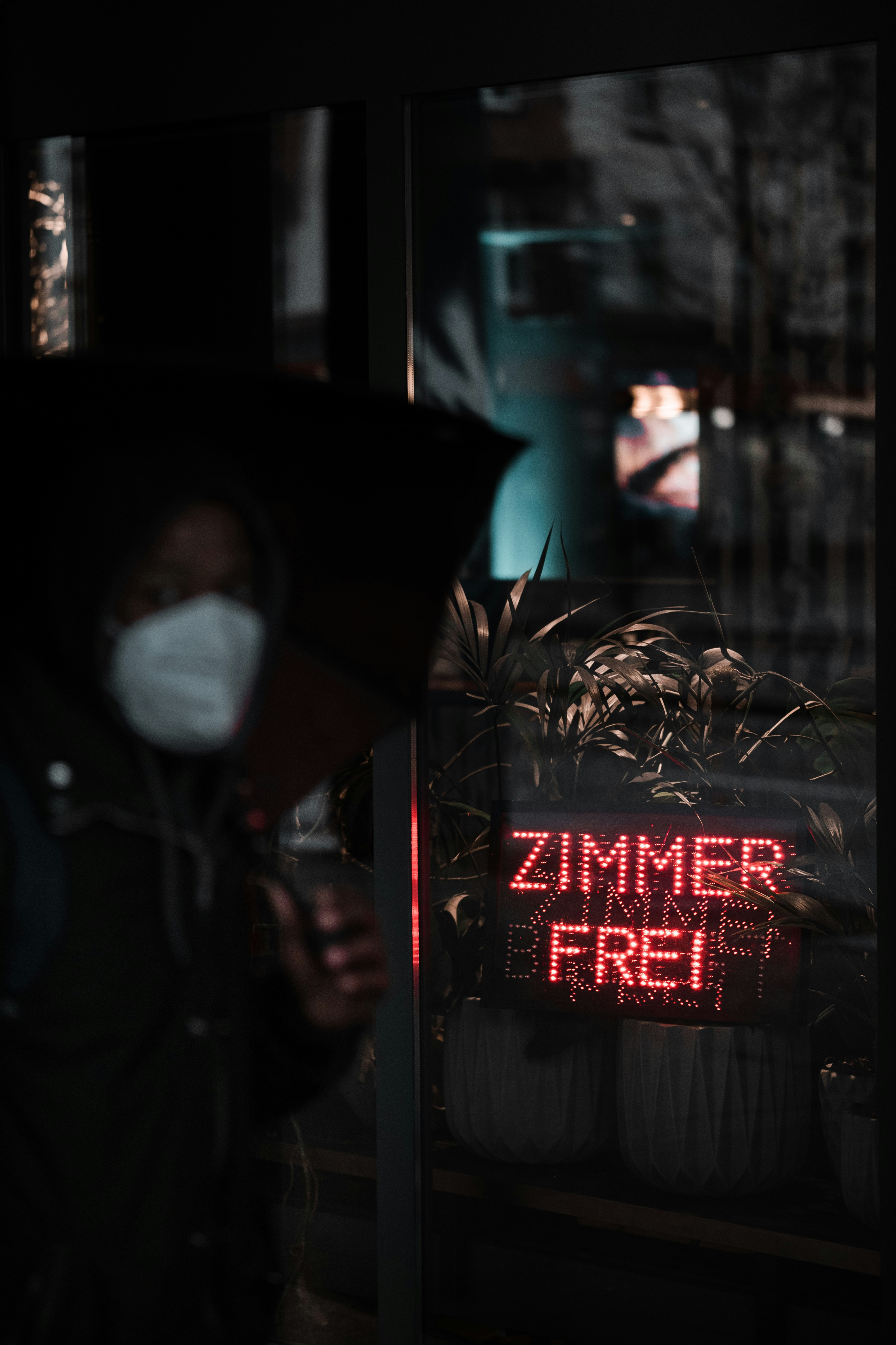 A person with an umbrella walks past a glowing 'ZIMMER FREI' sign, surrounded by indoor plants. The scene captures a blend of urban life and inviting warmth.