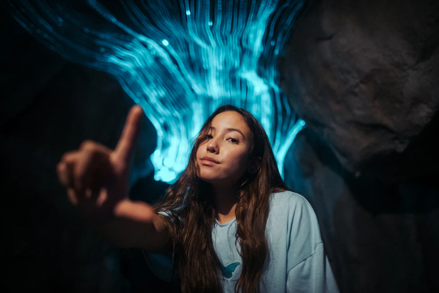 Woman posing in a dark setting with bright blue light streaks arcing behind her, her hand reaching toward the camera.