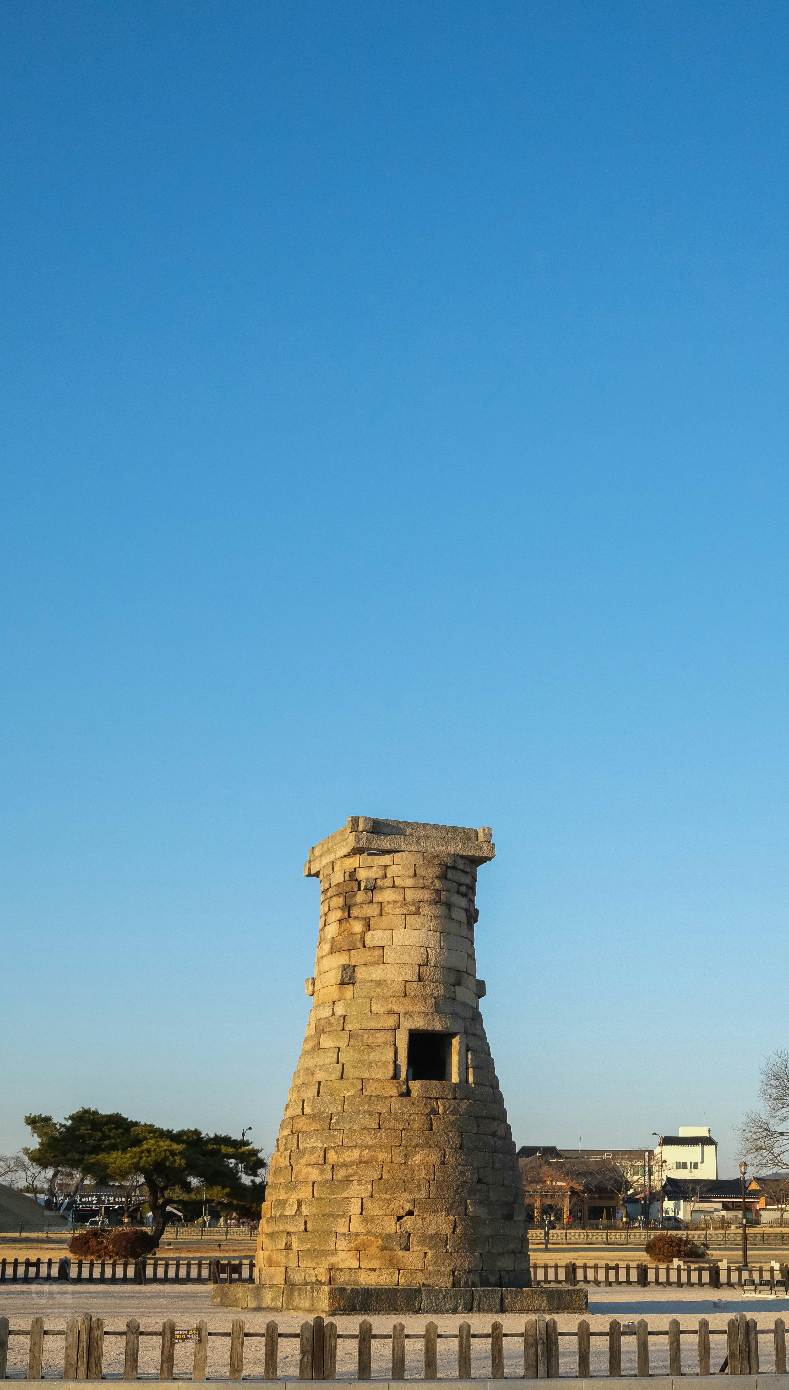 Historic stone tower standing against a clear blue sky, symbolizing ancient maritime navigation. The surrounding landscape features a wooden fence and sparse vegetation.