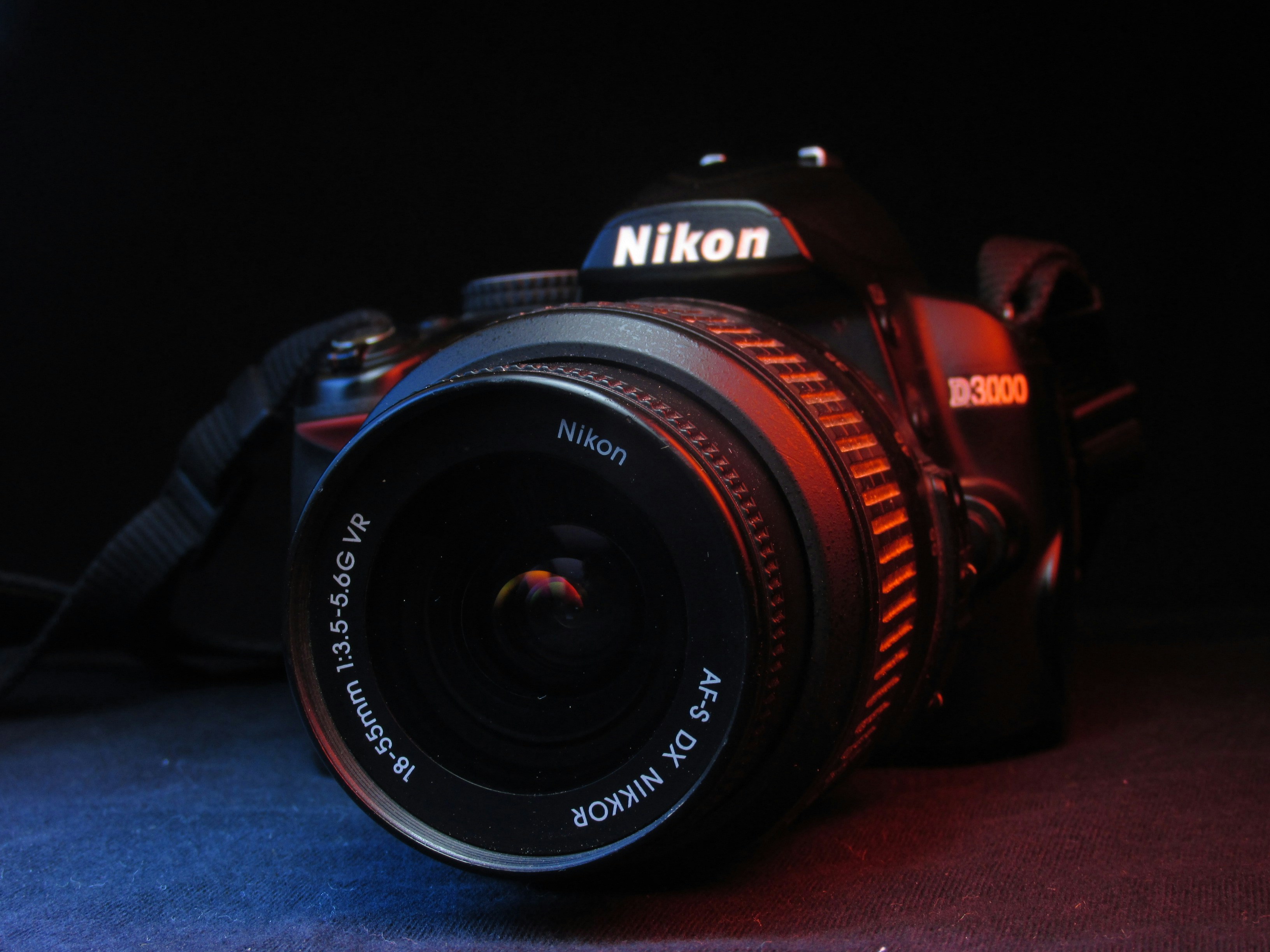 Nikon D3000 camera positioned on a dark surface, showcasing its lens and controls with subtle lighting effects.