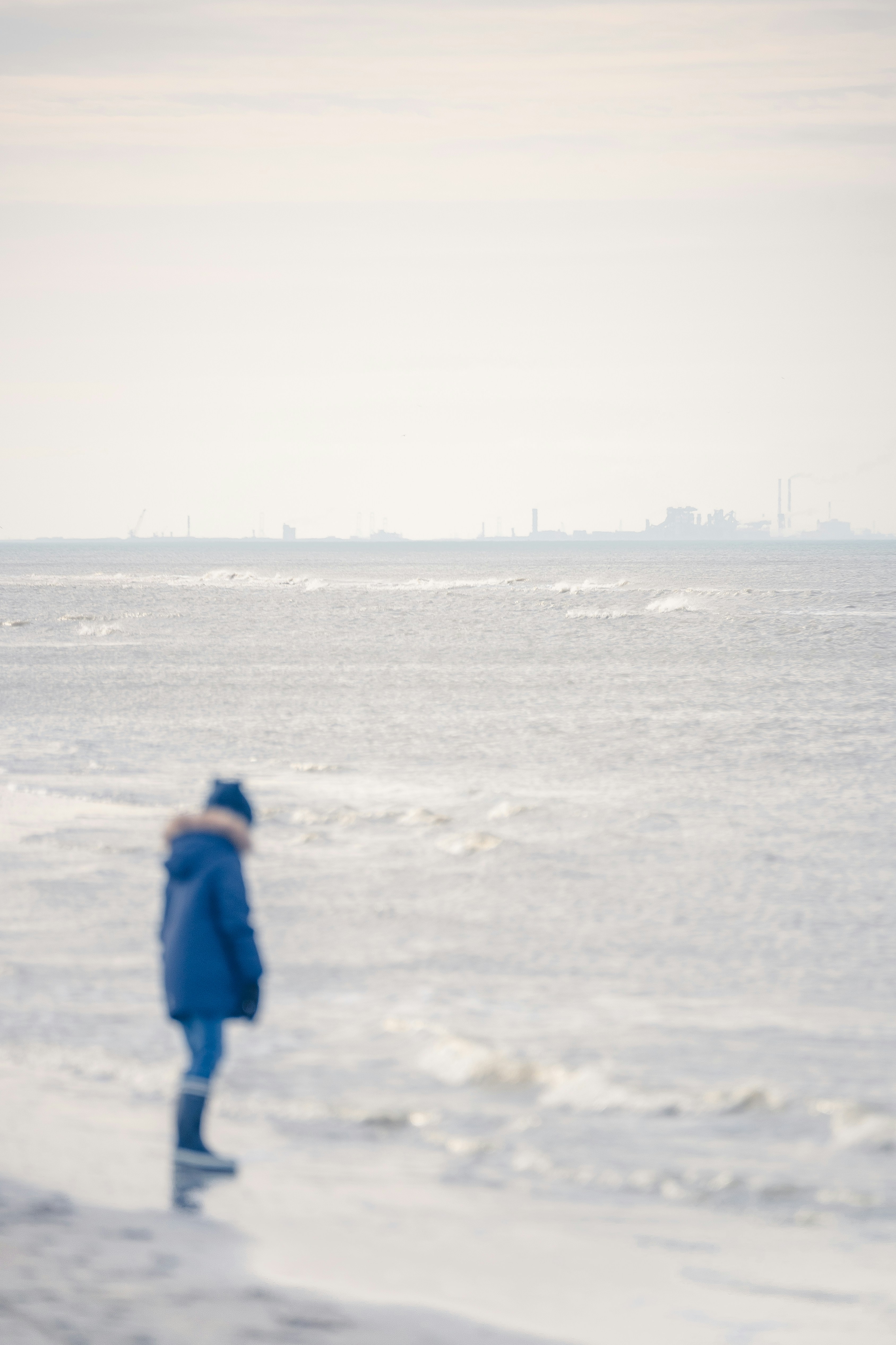 A figure in a blue coat stands at the water's edge, gazing out at the distant industrial skyline. The soft, muted colors create a serene atmosphere.
