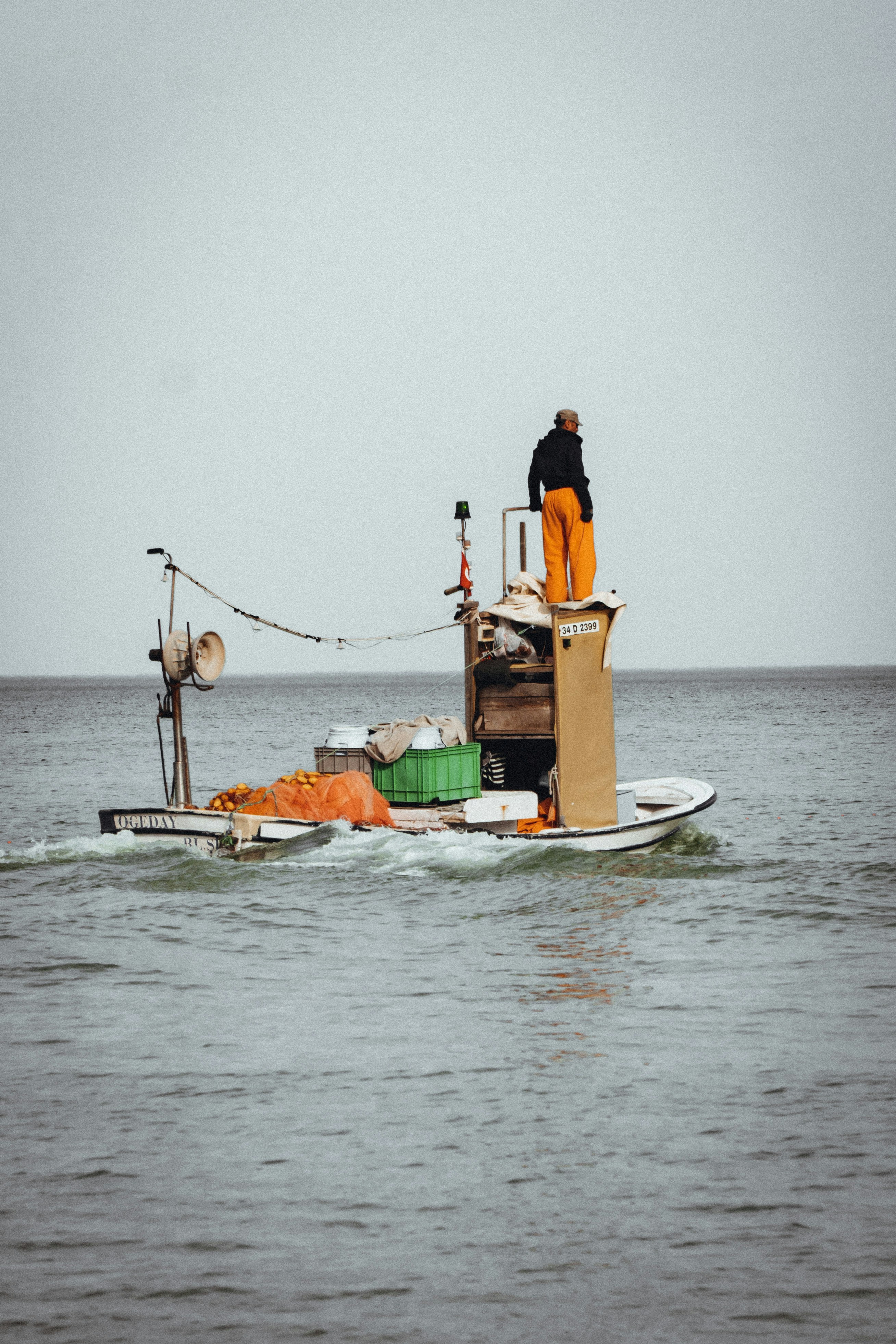 Tideward Vigil: Orange-Panted Figure on a Raised Platform Over a Cargo-Laden Boat