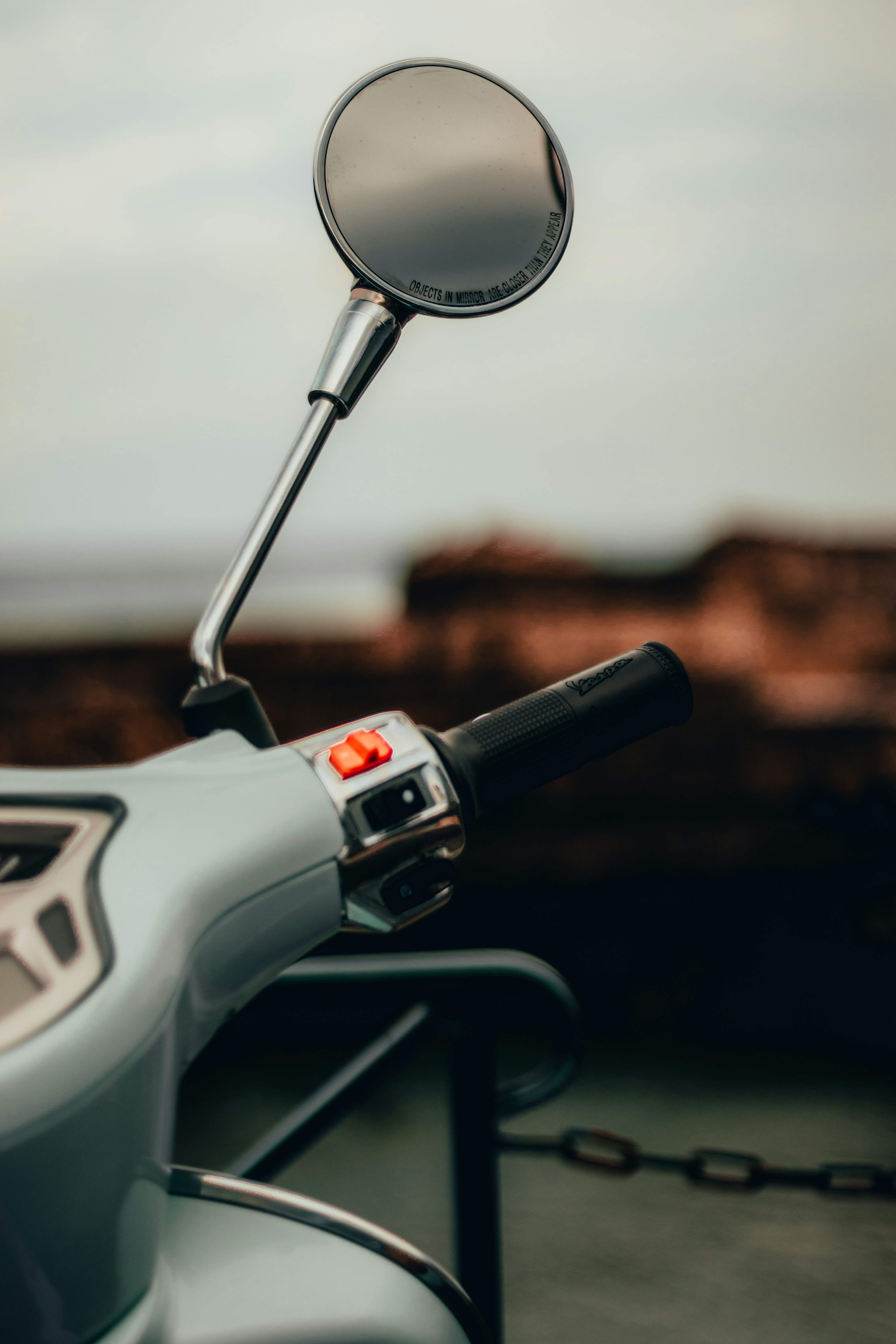 Close-up of a vintage scooter's handlebar and mirror, showcasing intricate details and textures. The blurred background hints at an adventurous journey ahead.