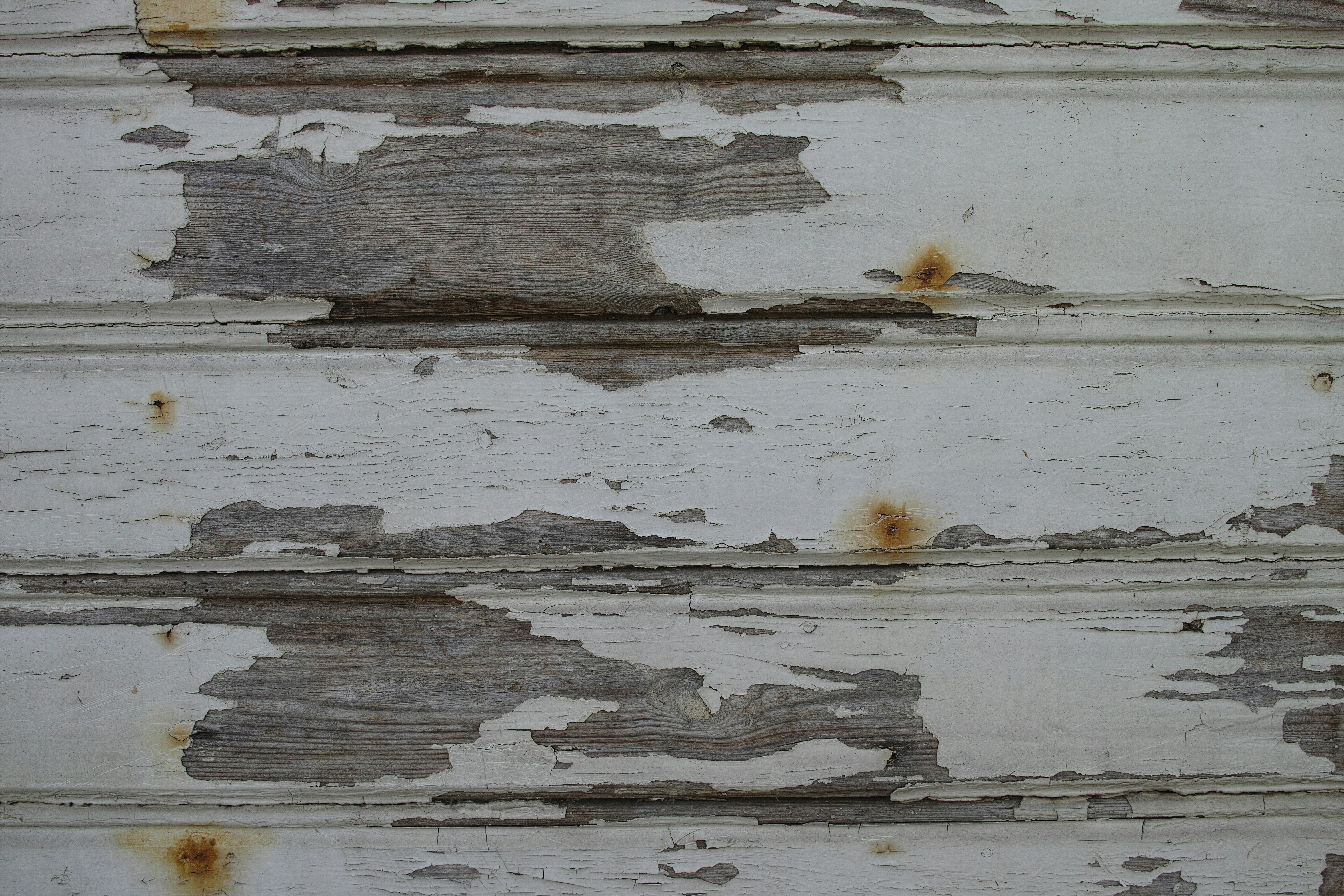 Close-up of peeling white paint on wooden planks, showcasing layers of texture and age. The surface reveals rust spots and natural wear.