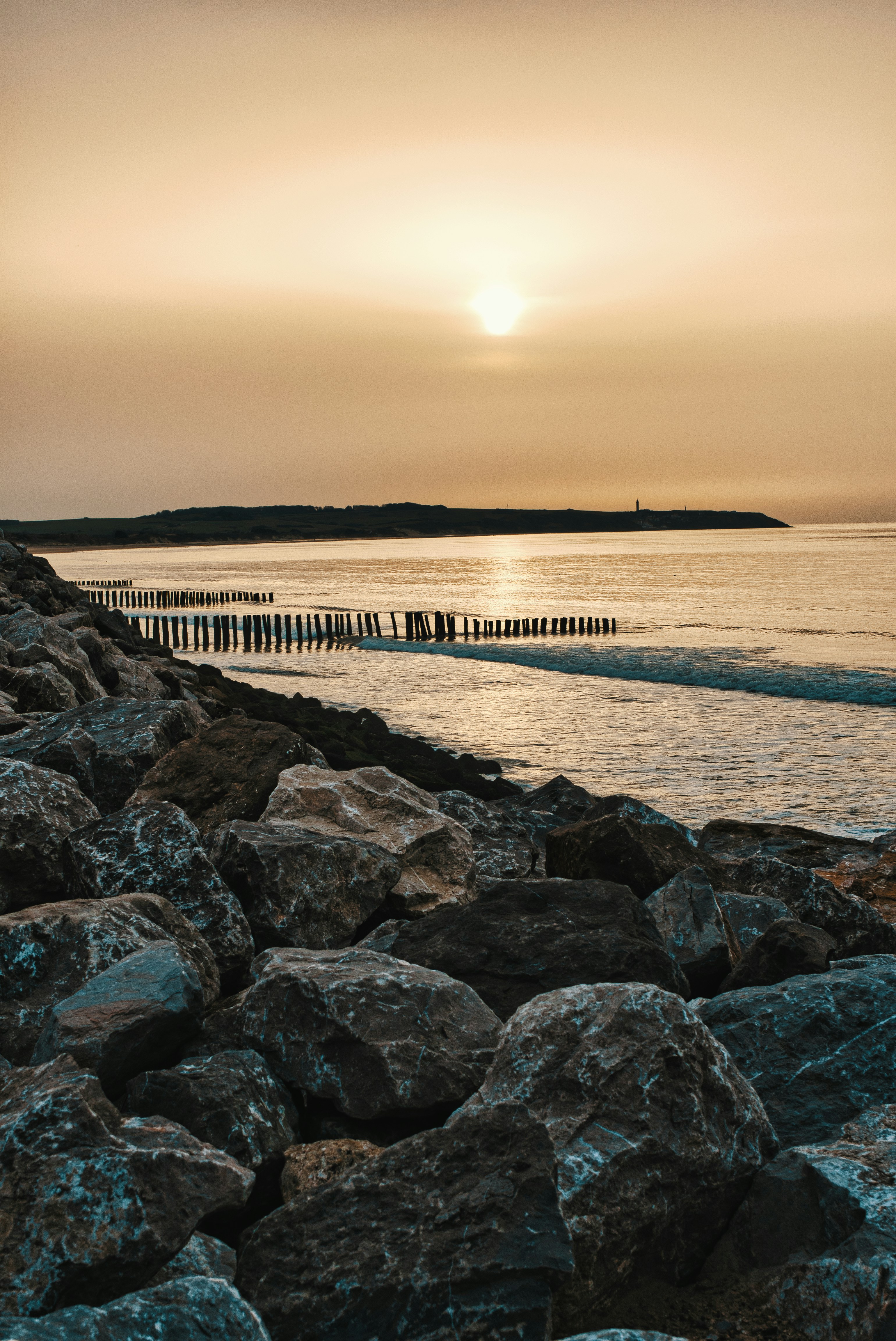 Golden sun setting over a calm sea, silhouetting weathered wooden posts along the shore, framed by rugged rocks in the foreground.