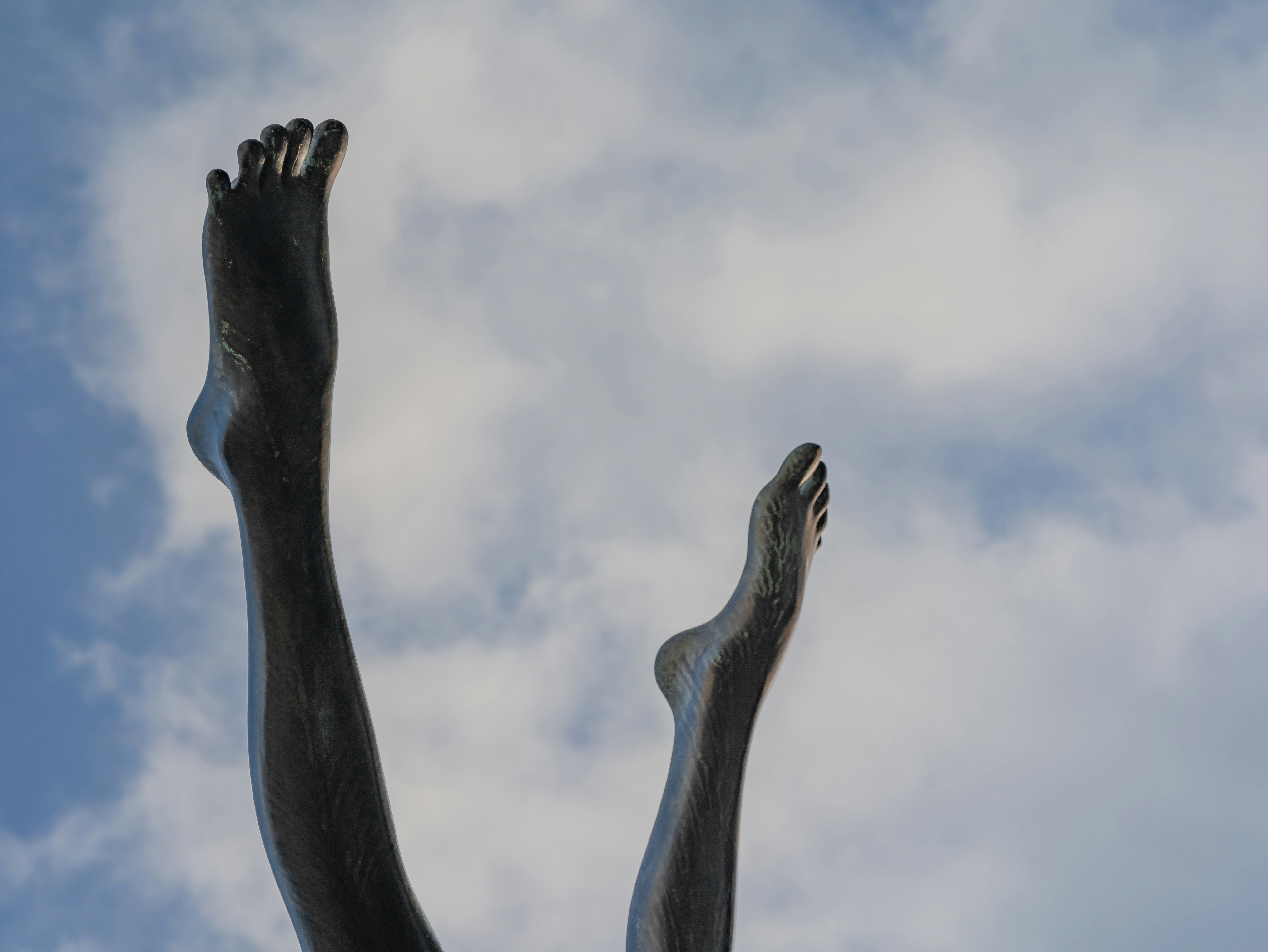 Bronze sculpture of two legs reaching upward against a backdrop of soft, cloudy sky.