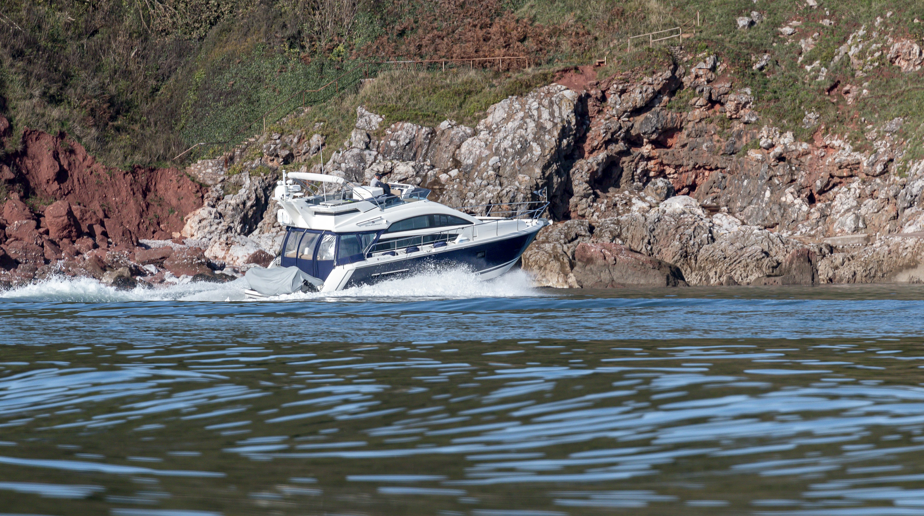 Motor yacht glides swiftly near rugged cliffs, leaving a wake in calm coastal waters.