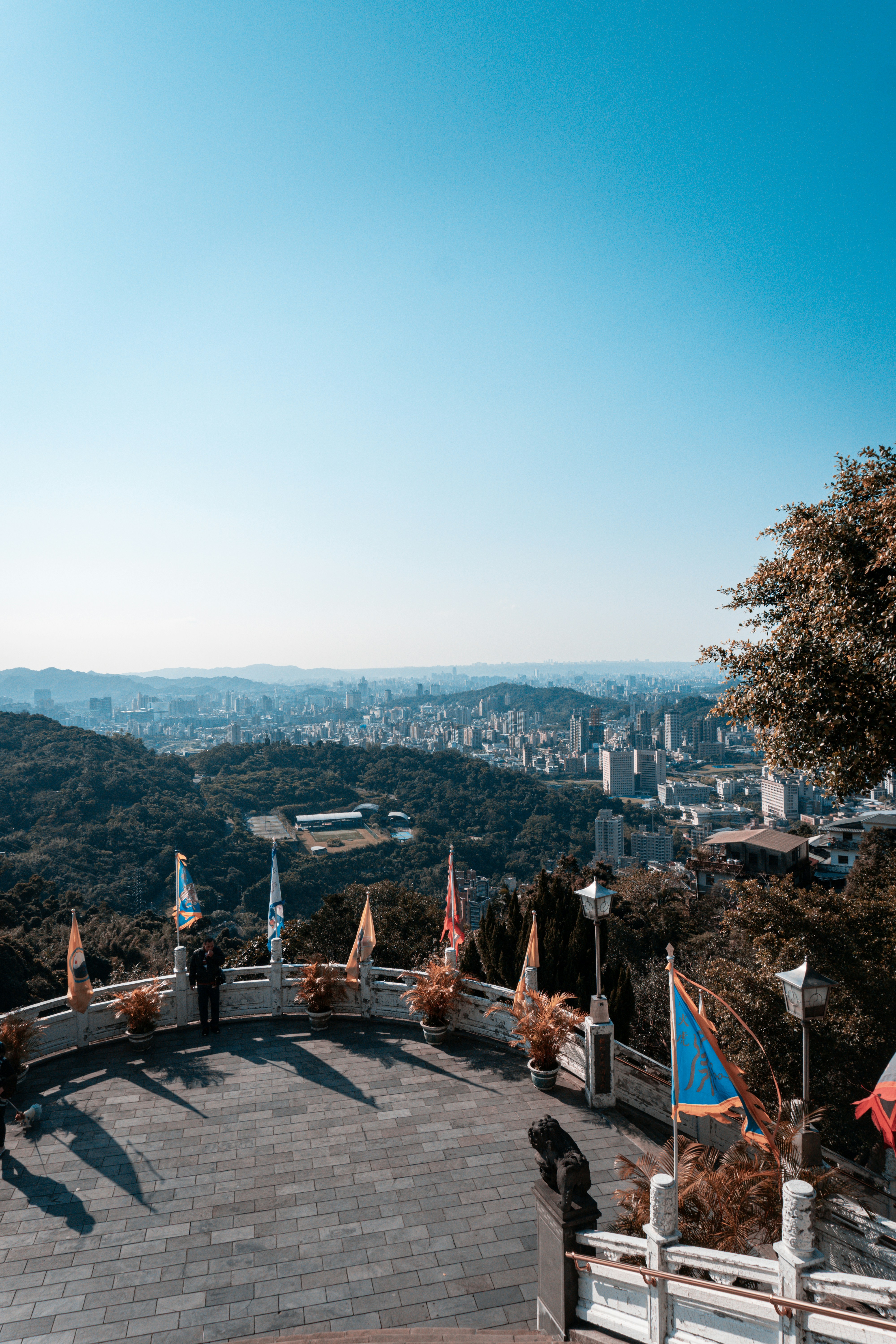 Visitors admire a panoramic view from a scenic overlook, with cityscape and mountains in the background. Colorful flags flutter in the gentle breeze.