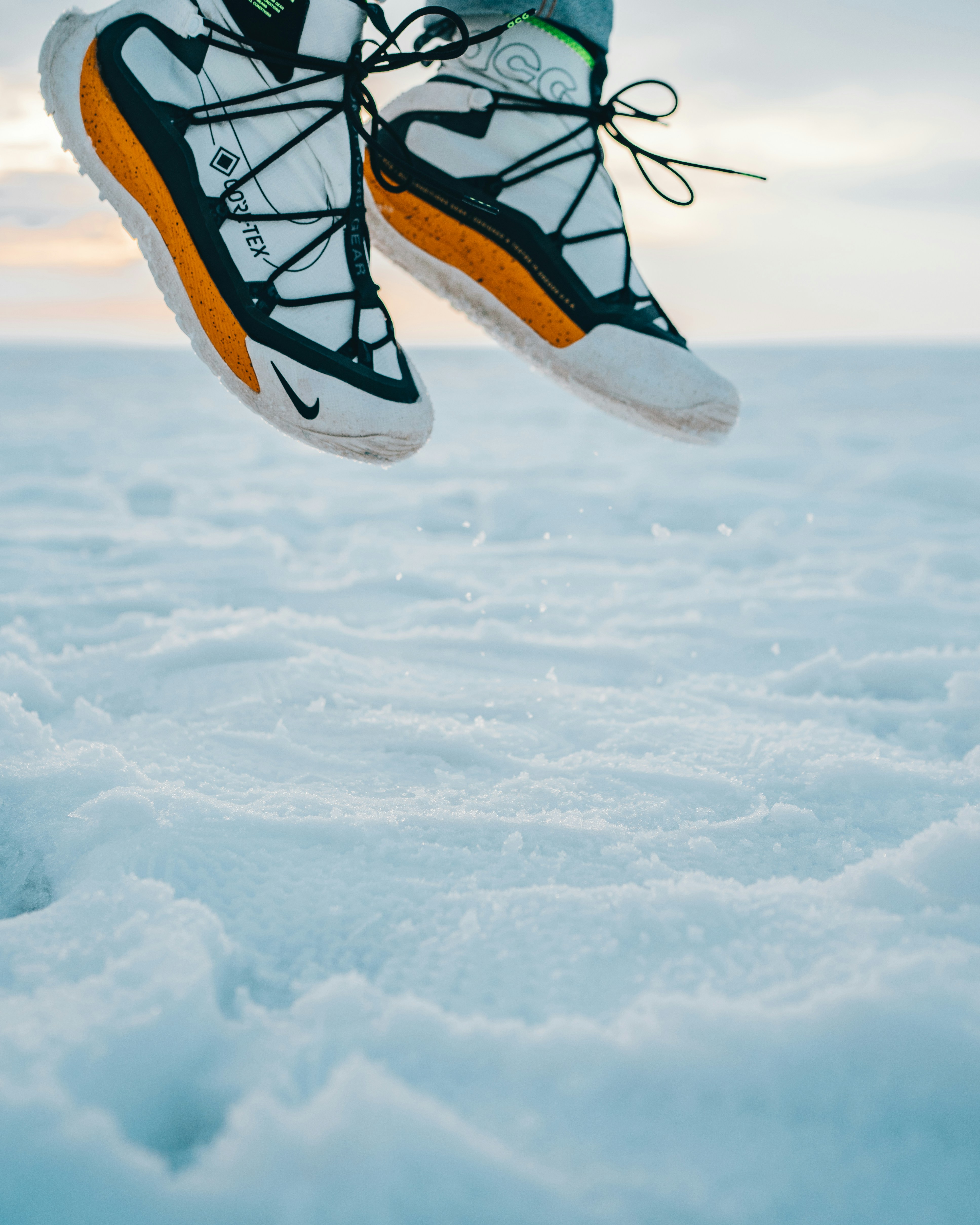 Neon-accented sneakers hover mid-air above a snow-covered landscape, captured in a crisp, action-filled moment. The shot emphasizes motion and the texture of powdery snow.