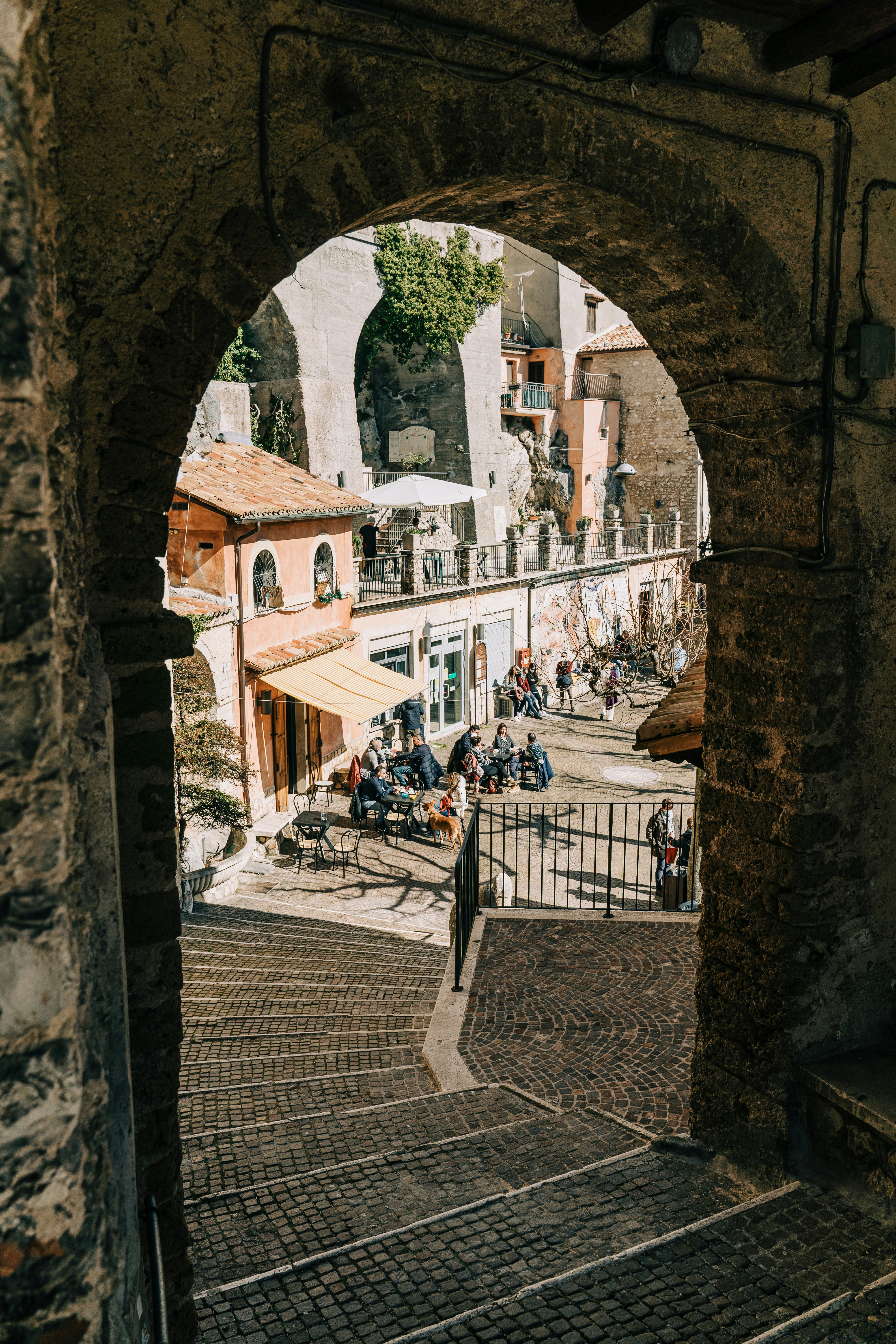 Charming courtyard scene framed by an ancient archway, showcasing lively gatherings and vibrant architecture. Sunlight casts warm tones across cobblestone paths.