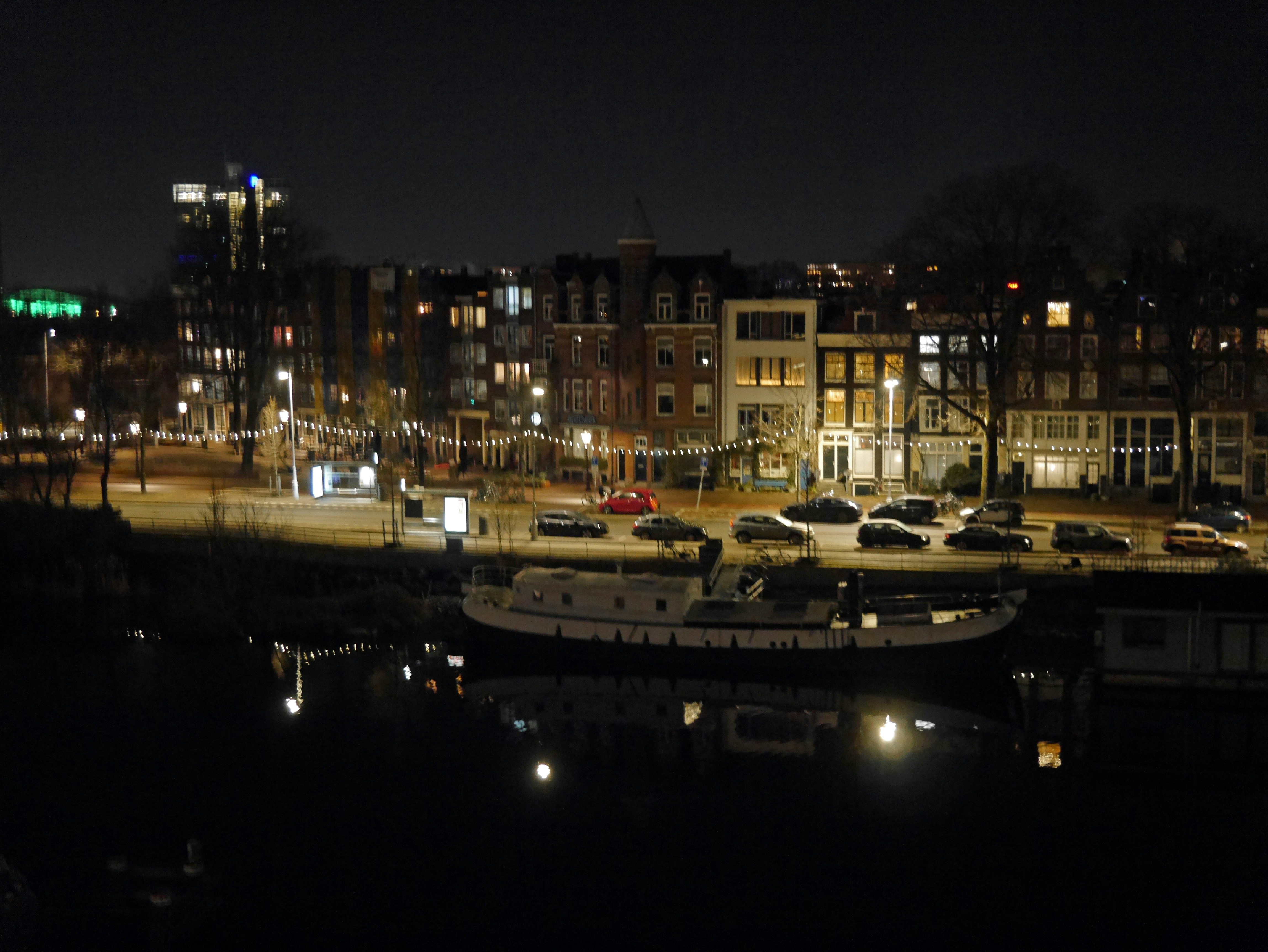 Cityscape at night featuring illuminated buildings along a waterfront, with reflections shimmering on the water's surface.