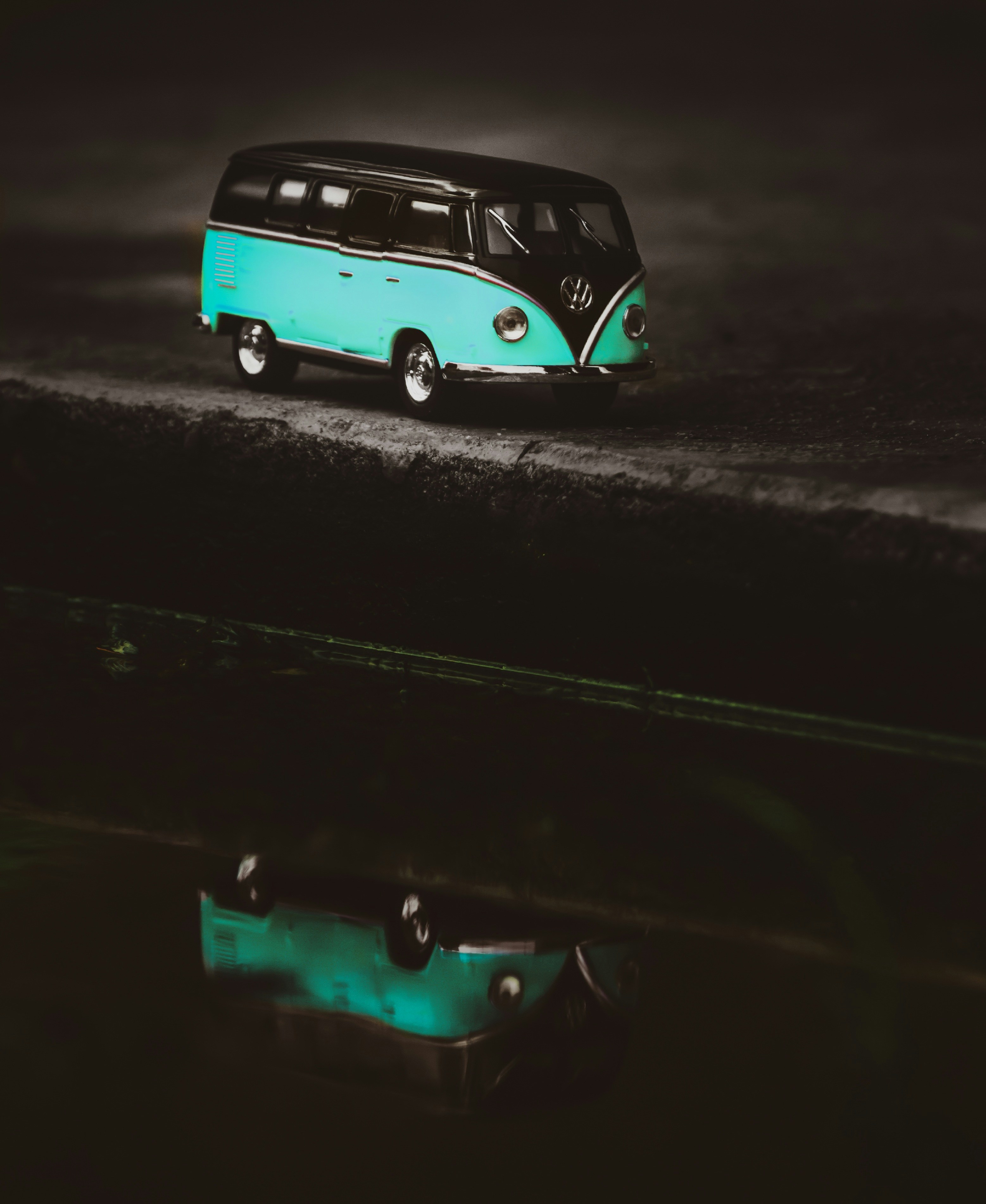 A vintage turquoise VW bus model perched on the edge of a reflective surface, evoking nostalgia and adventure.