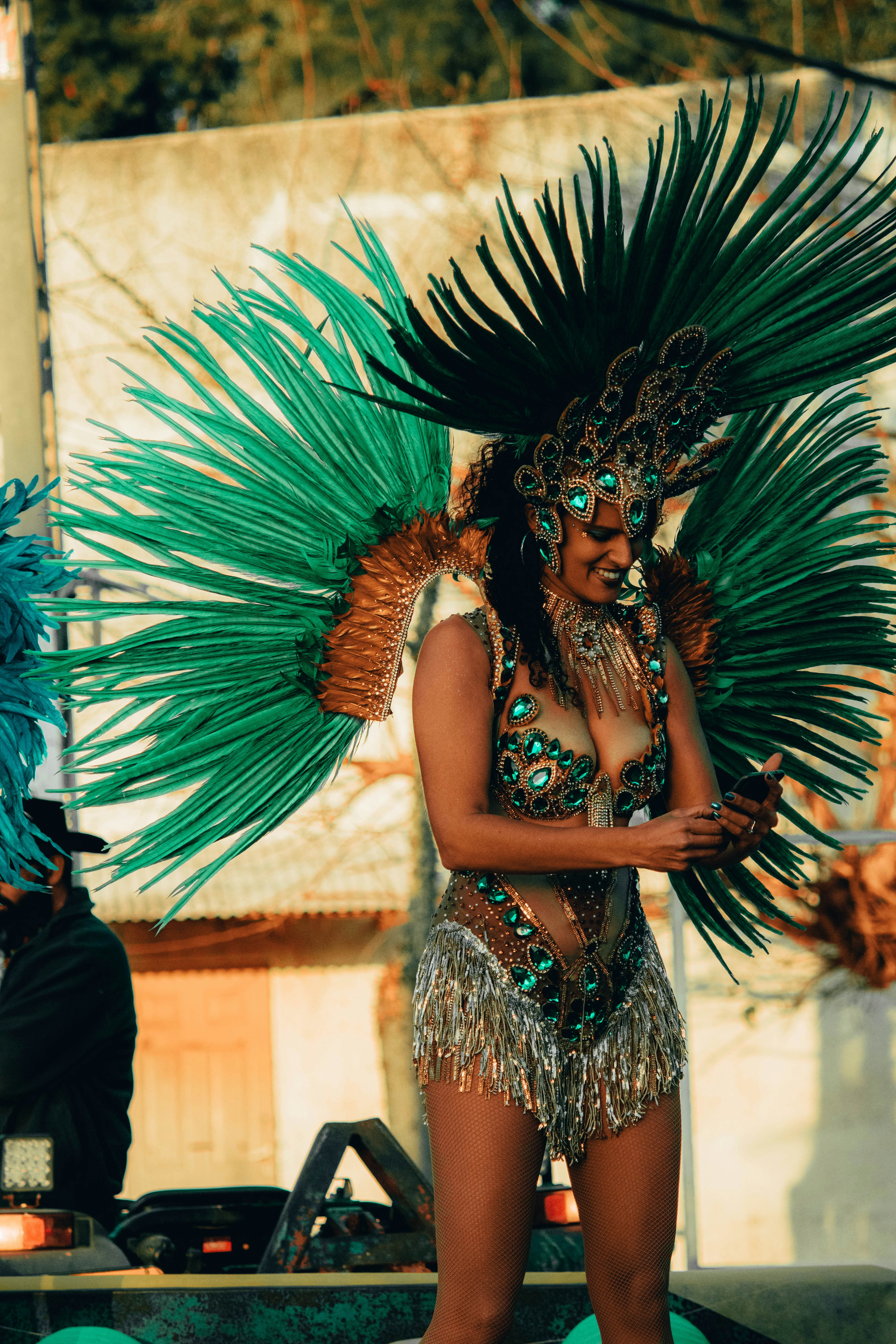 A performer adorned in a vibrant costume with elaborate feathered accessories interacts with her phone during a festive celebration.