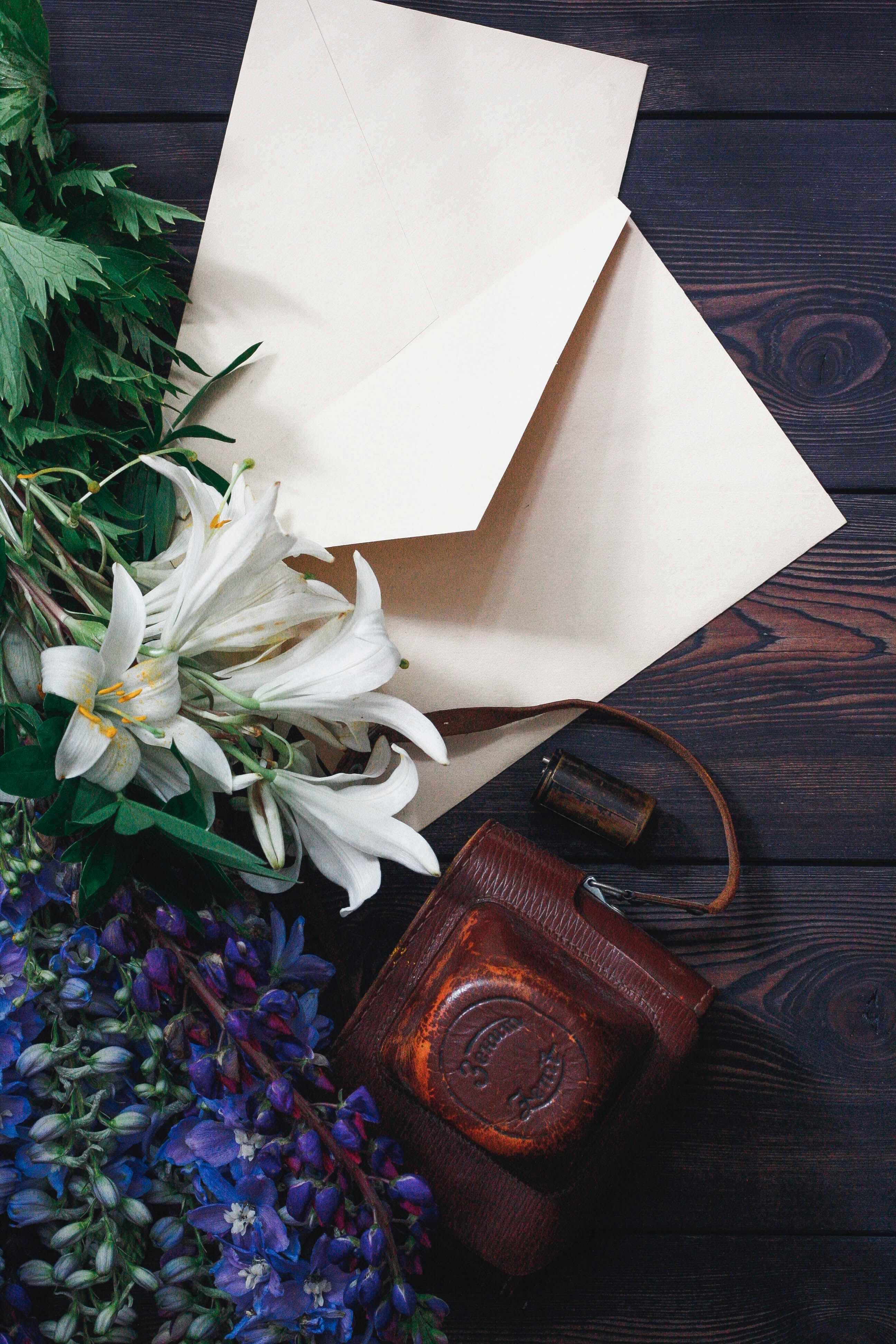 A curated arrangement of white lilies, blue flowers, and a leather pouch, accompanied by blank envelopes on a wooden surface.