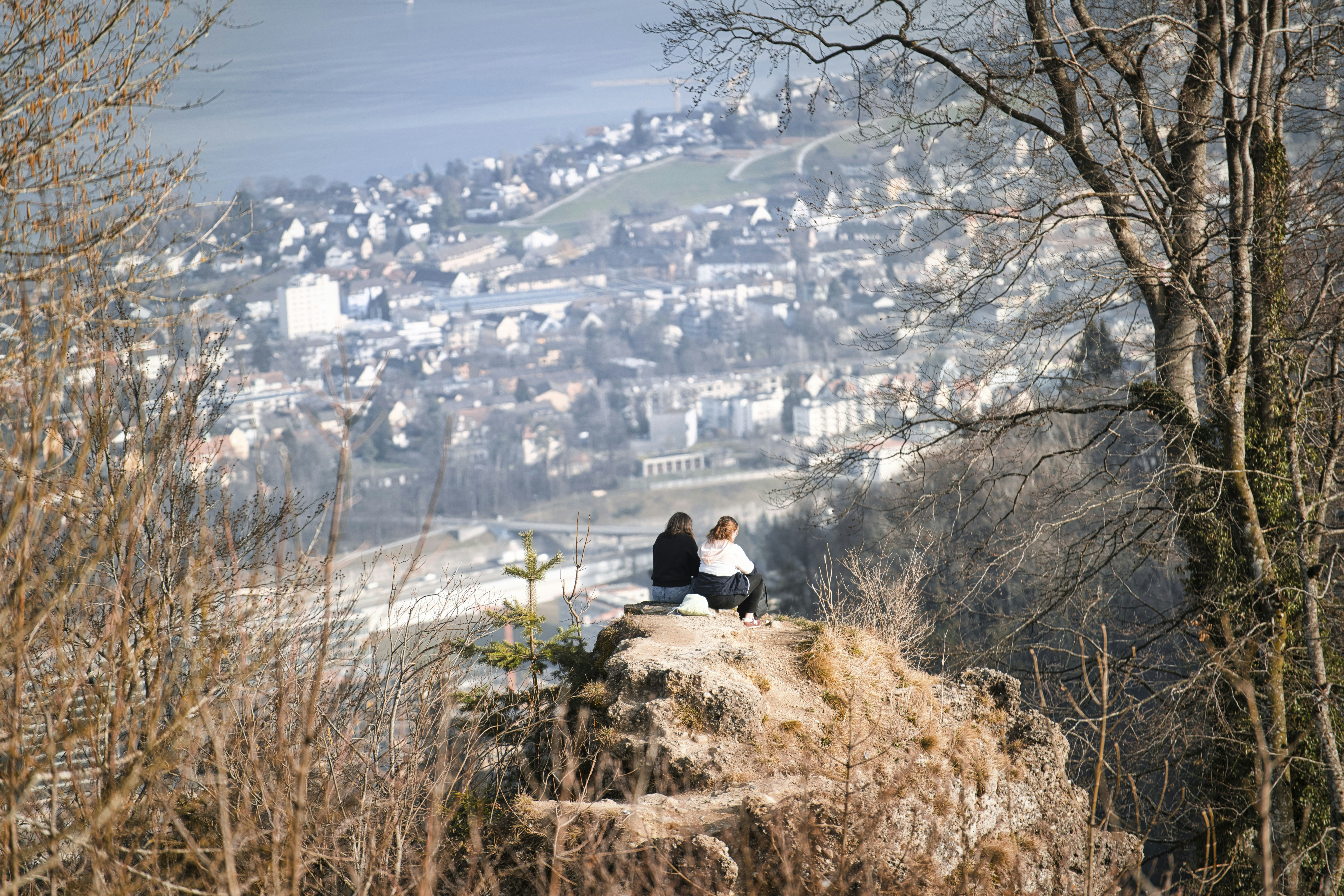 Two individuals sitting on a rocky outcrop overlooking a sprawling valley and distant hills. The scene captures a moment of tranquility amidst nature.