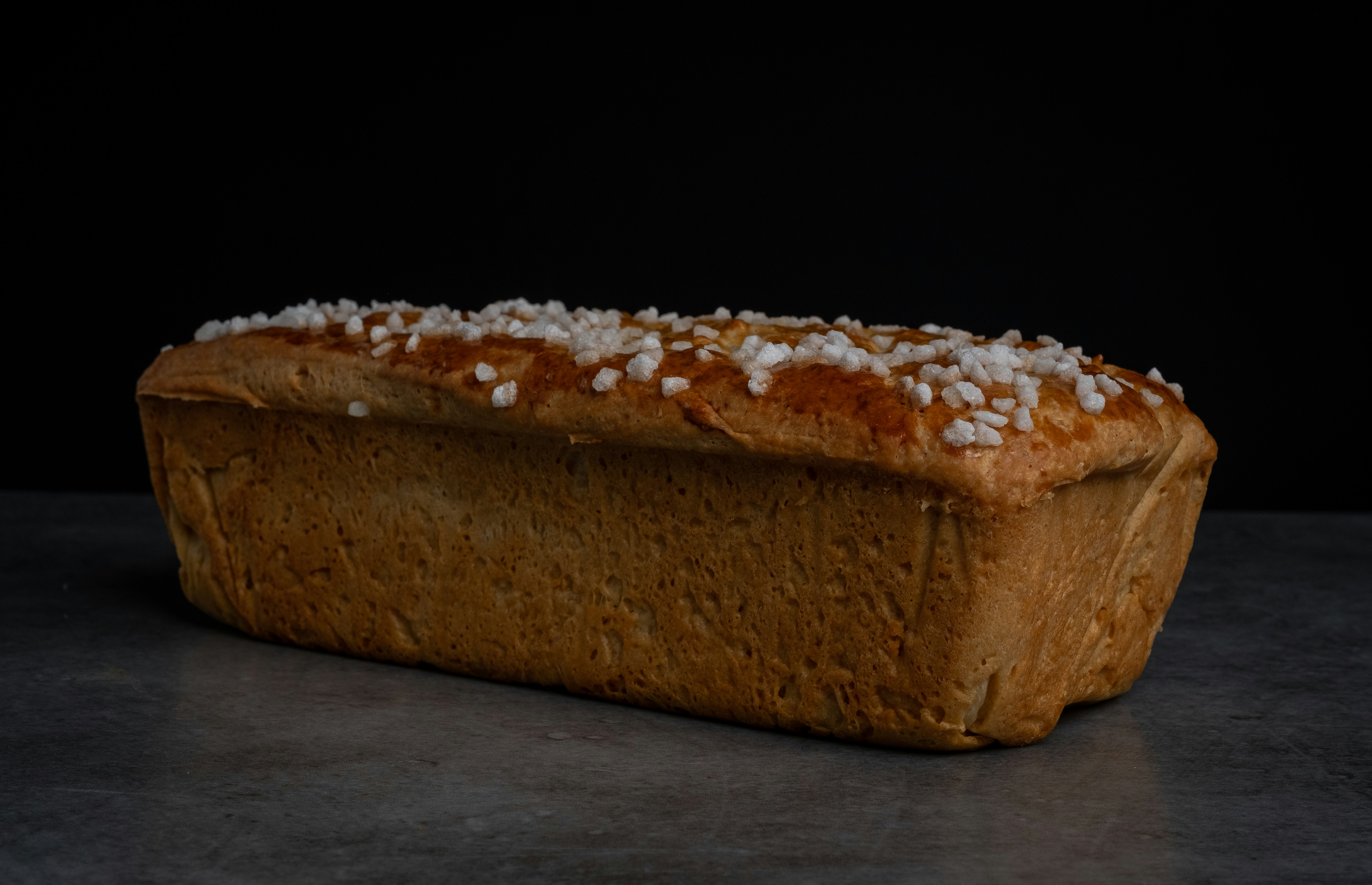 Freshly baked loaf of bread topped with coarse salt, resting on a textured surface against a dark background.