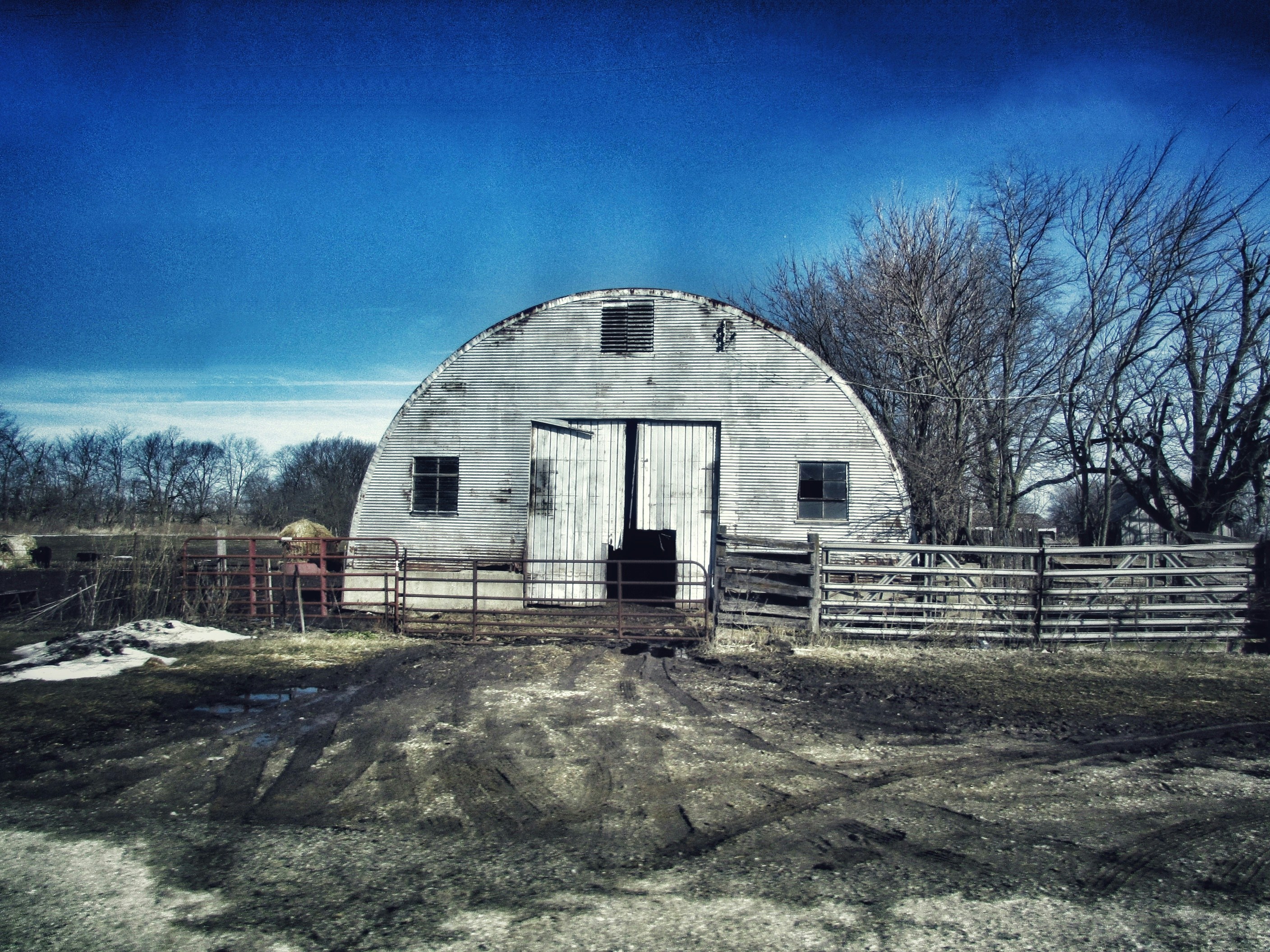 Weathered round metal barn sits behind a wooden corral in a muddy farmyard under a bright blue sky. This is a straightforward rural photograph.