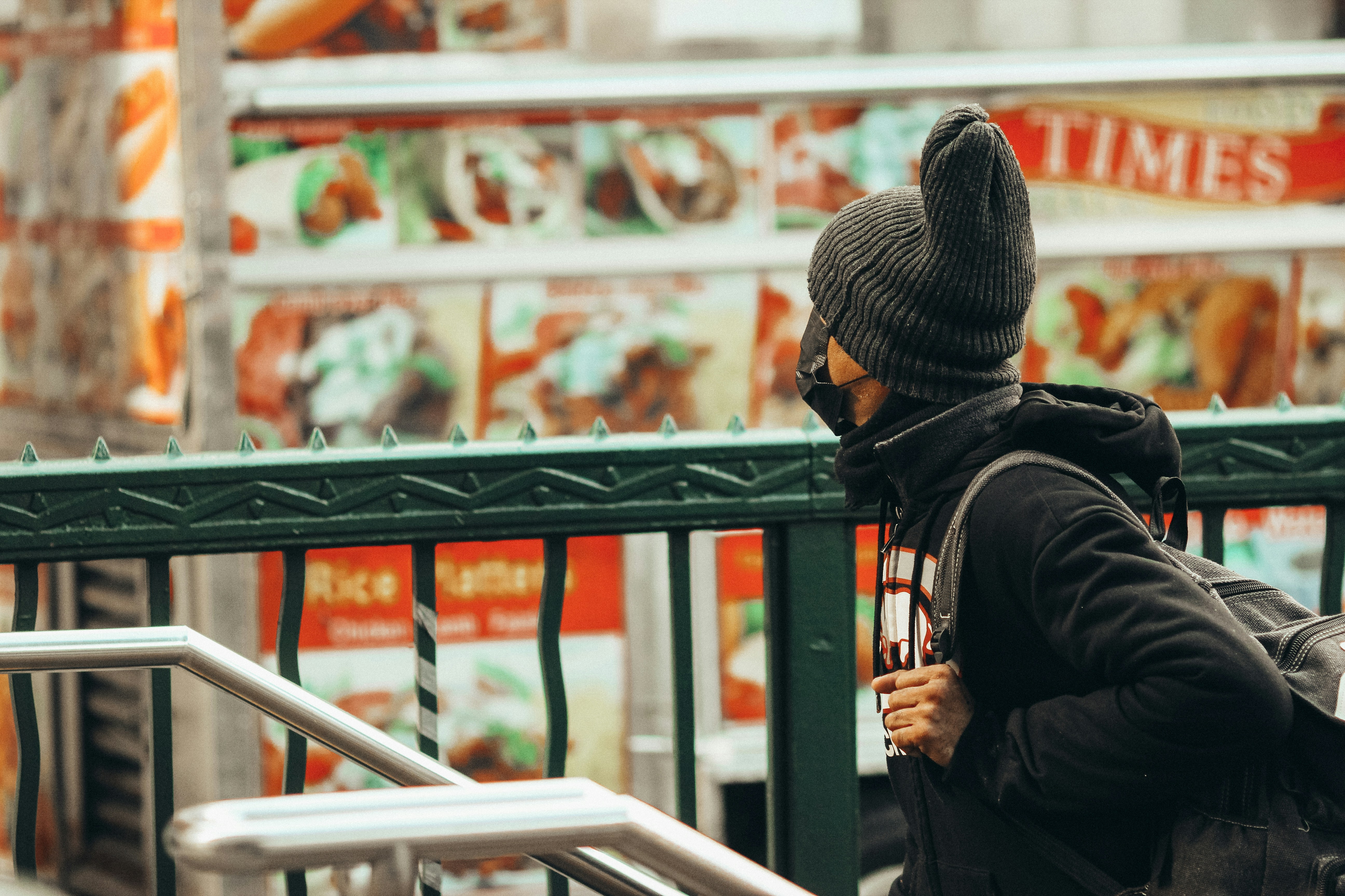 Individual in a beanie and mask walks past vibrant food advertisements on a city street.
