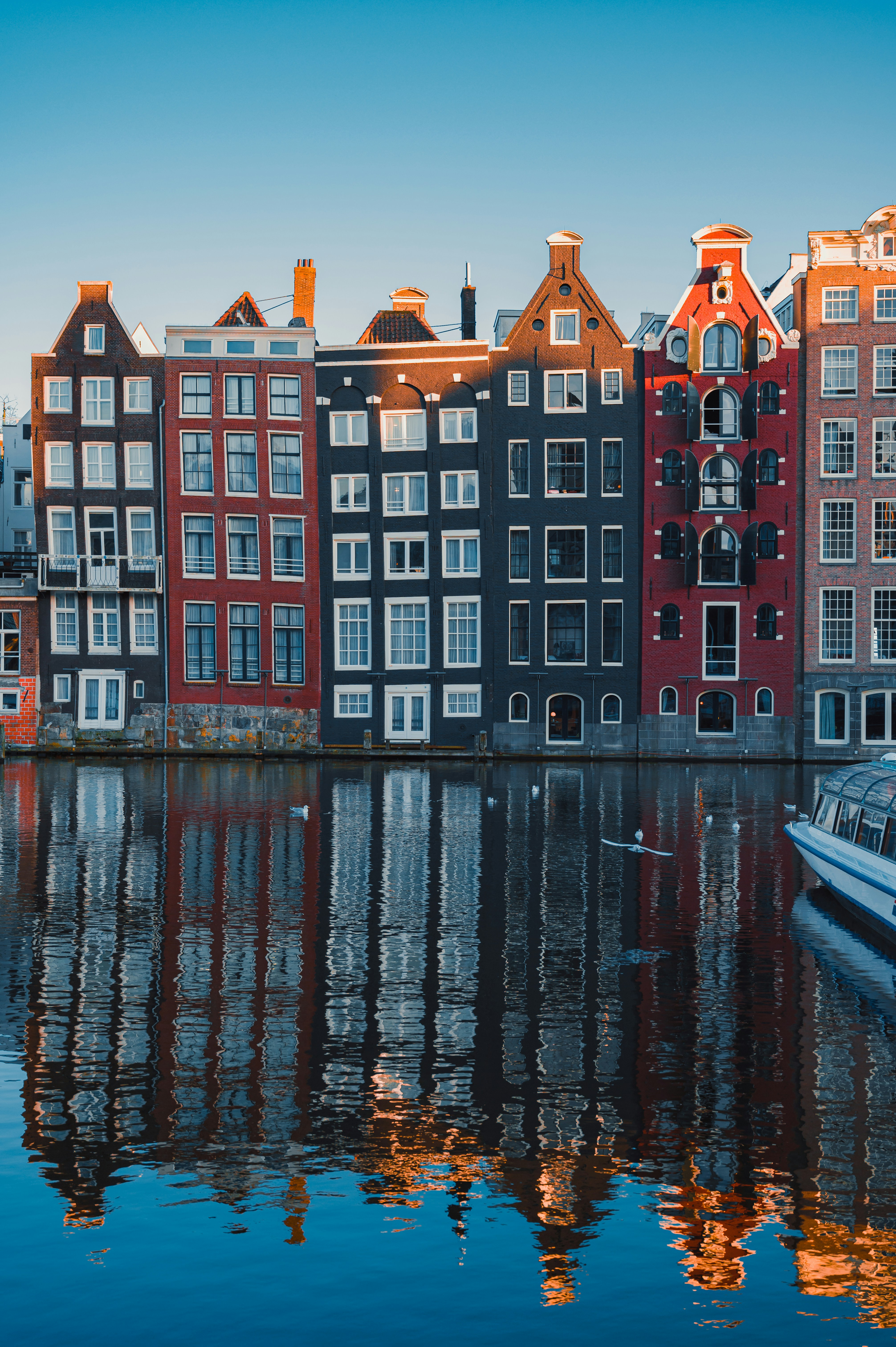 Vibrant canal houses along the water's edge in Amsterdam, showcasing a mix of architectural styles and colors. The reflection in the calm water enhances the visual appeal.