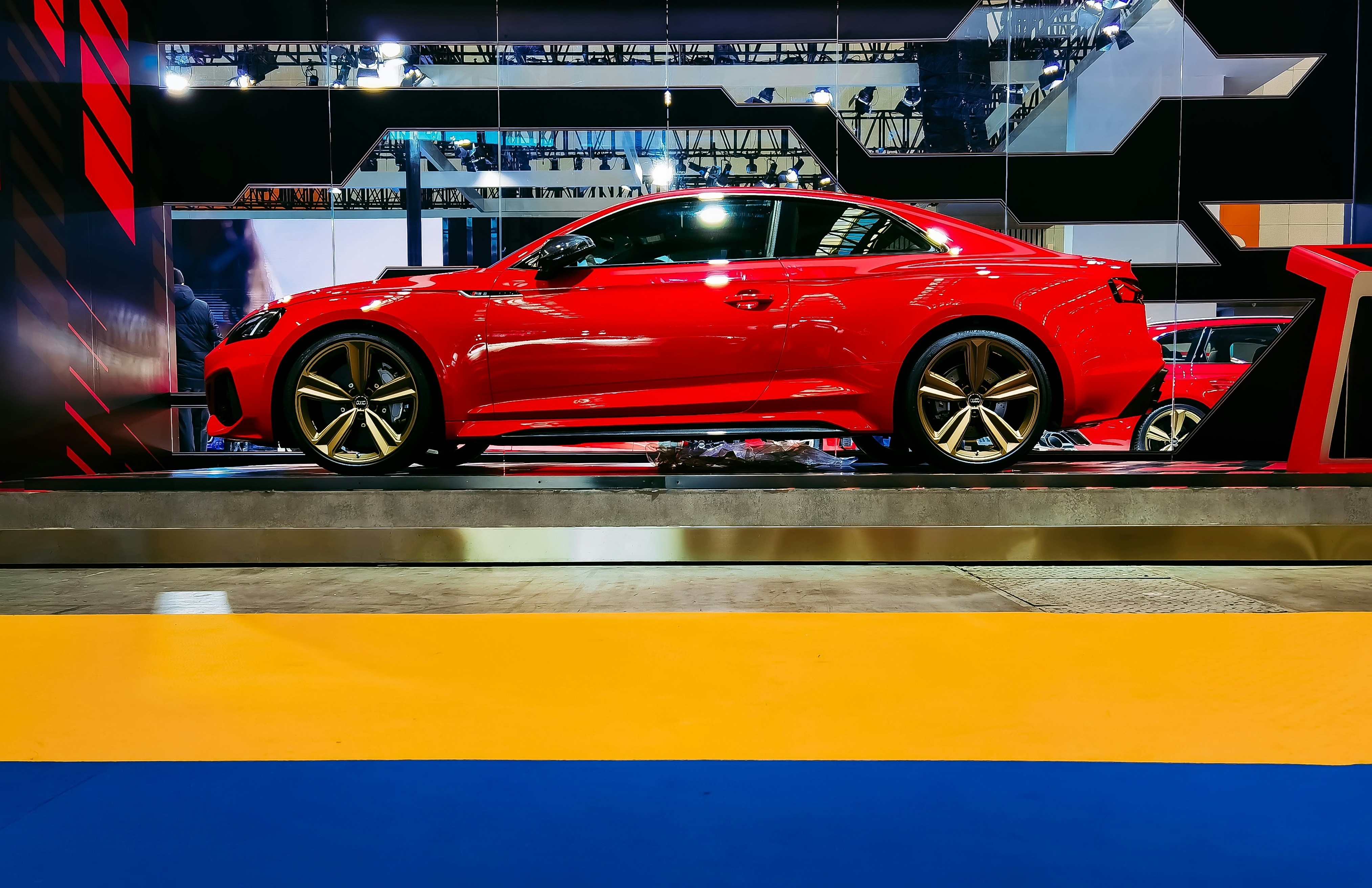 Red sports coupe on a glossy showroom platform under bright exhibition lighting.