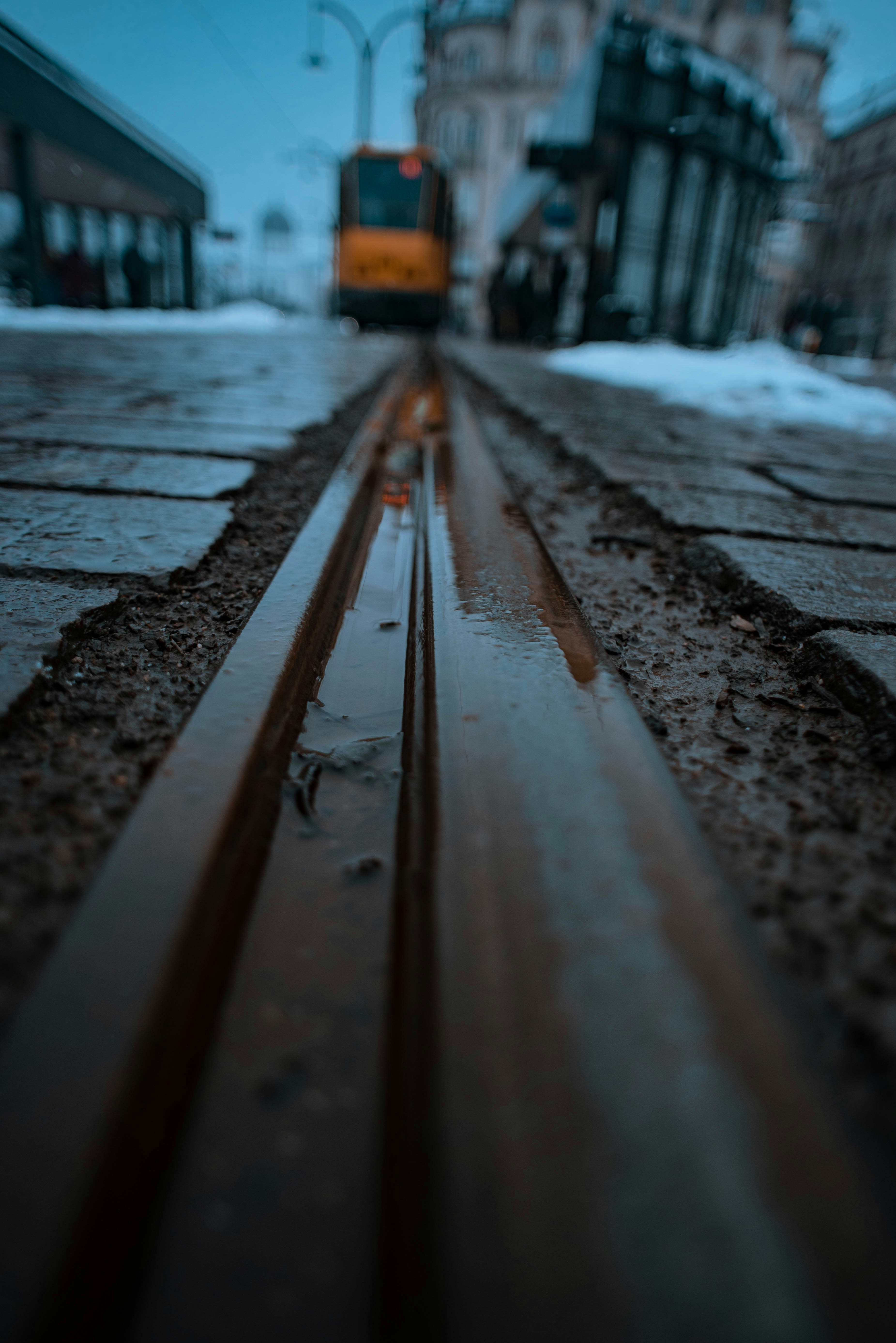 Tram tracks glisten with rainwater, leading towards a blurred tram in a bustling cityscape. Snow-covered surroundings add a touch of winter's chill.