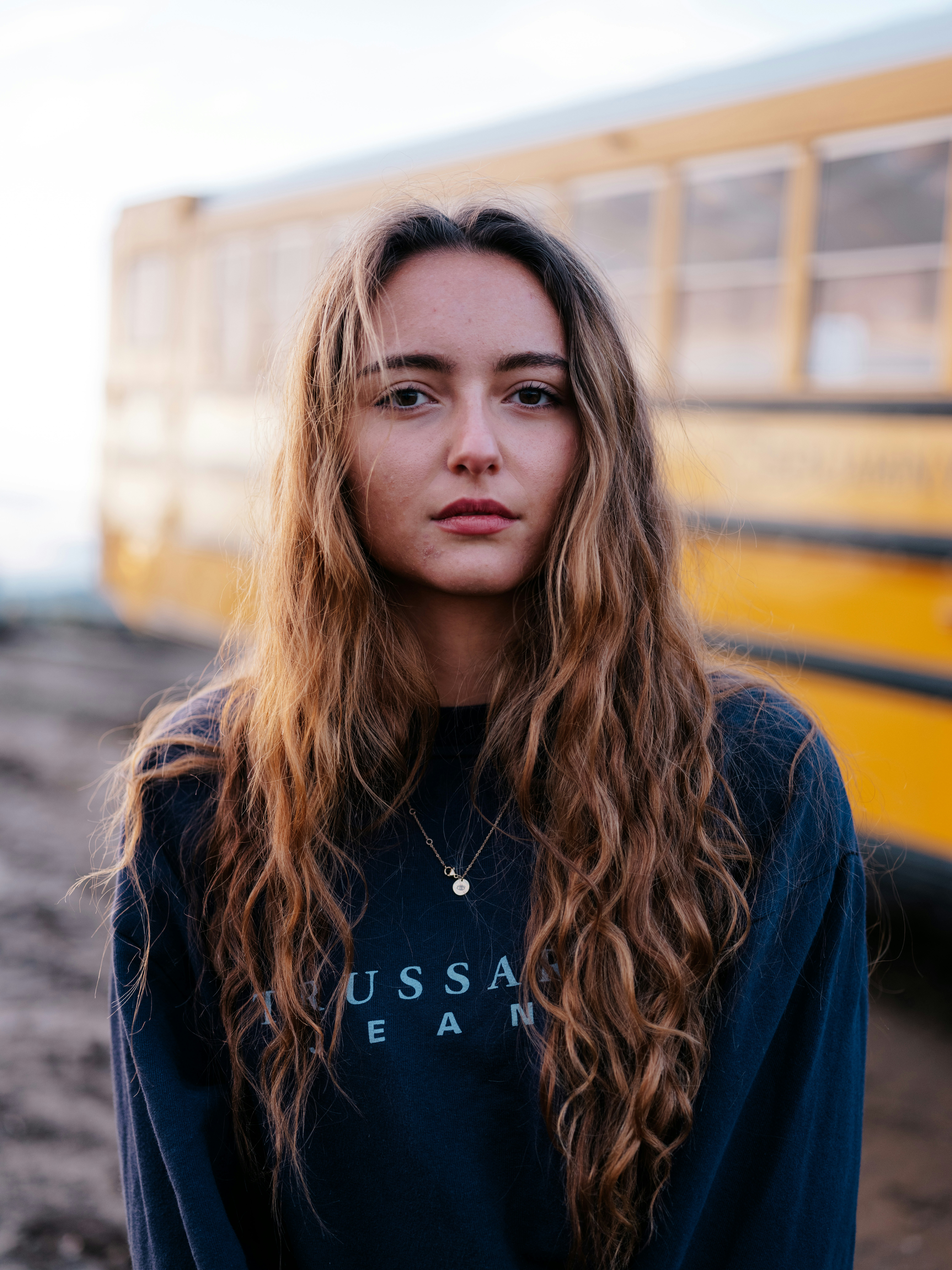 Young woman with long, wavy hair gazes thoughtfully at the camera, set against a blurred yellow school bus in the background.