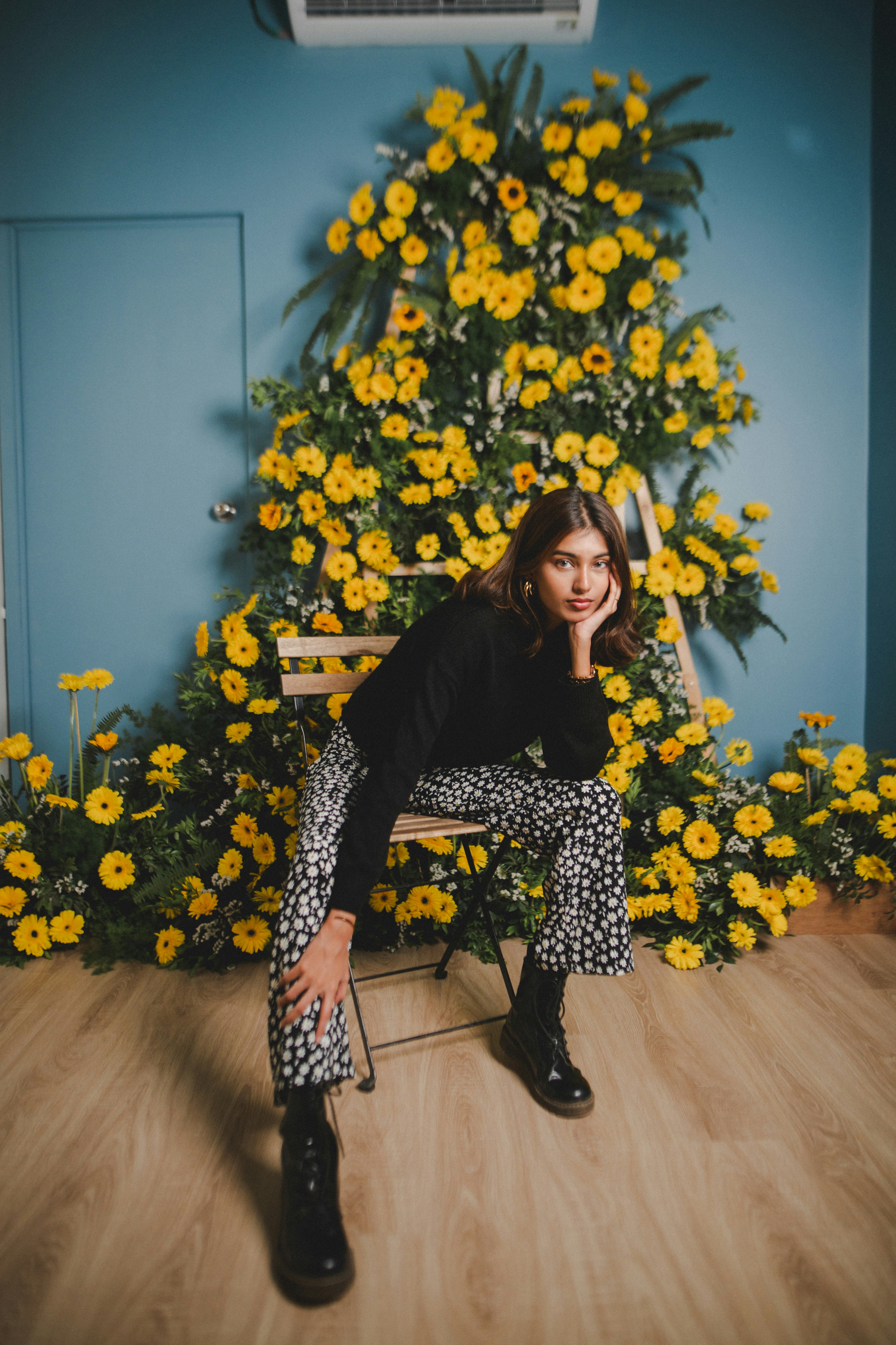 Model posed thoughtfully in front of a vibrant wall of yellow flowers, showcasing a stylish outfit with patterned pants and a black top.
