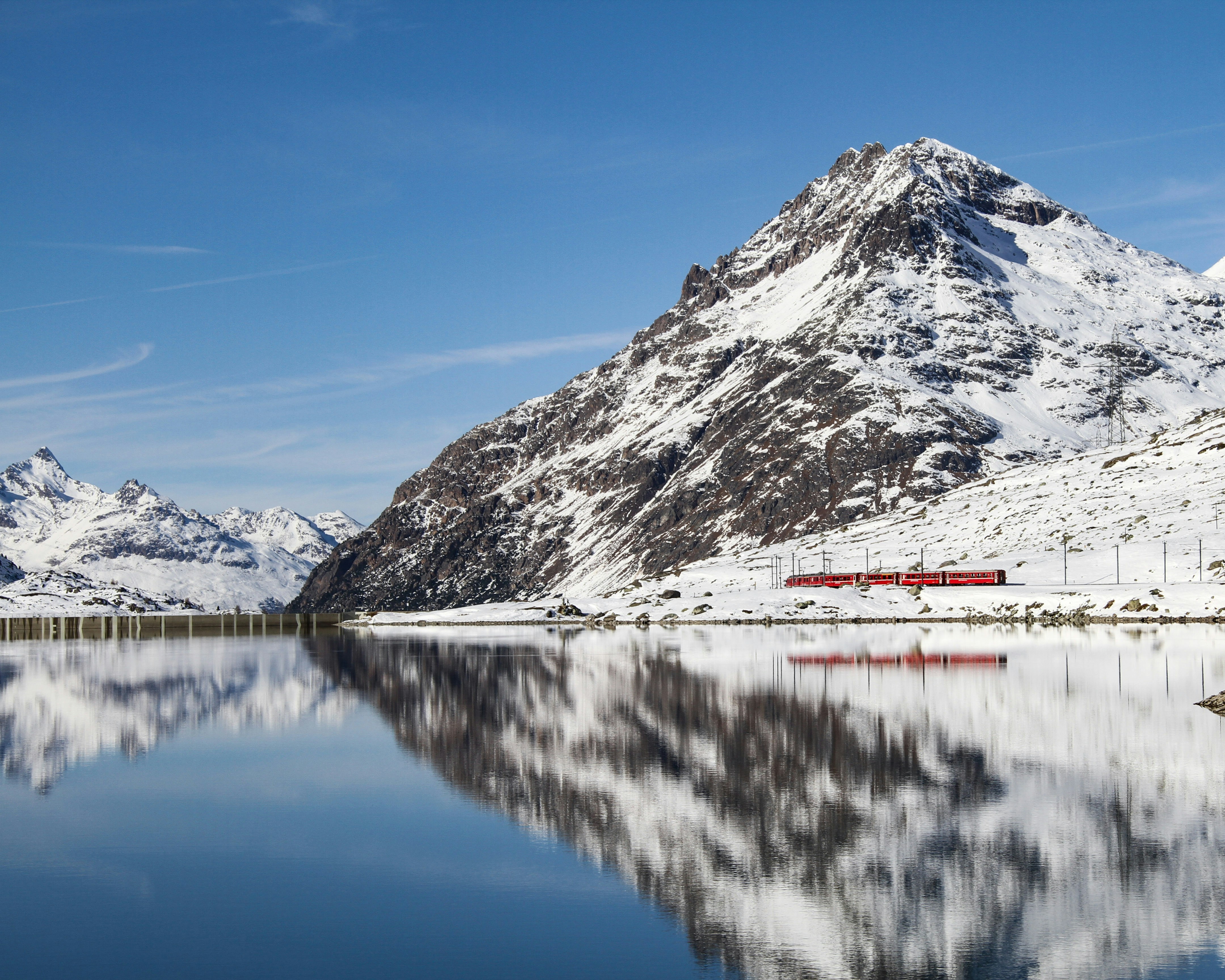 Snow-capped mountain reflecting on a tranquil lake with a red train traversing the landscape.